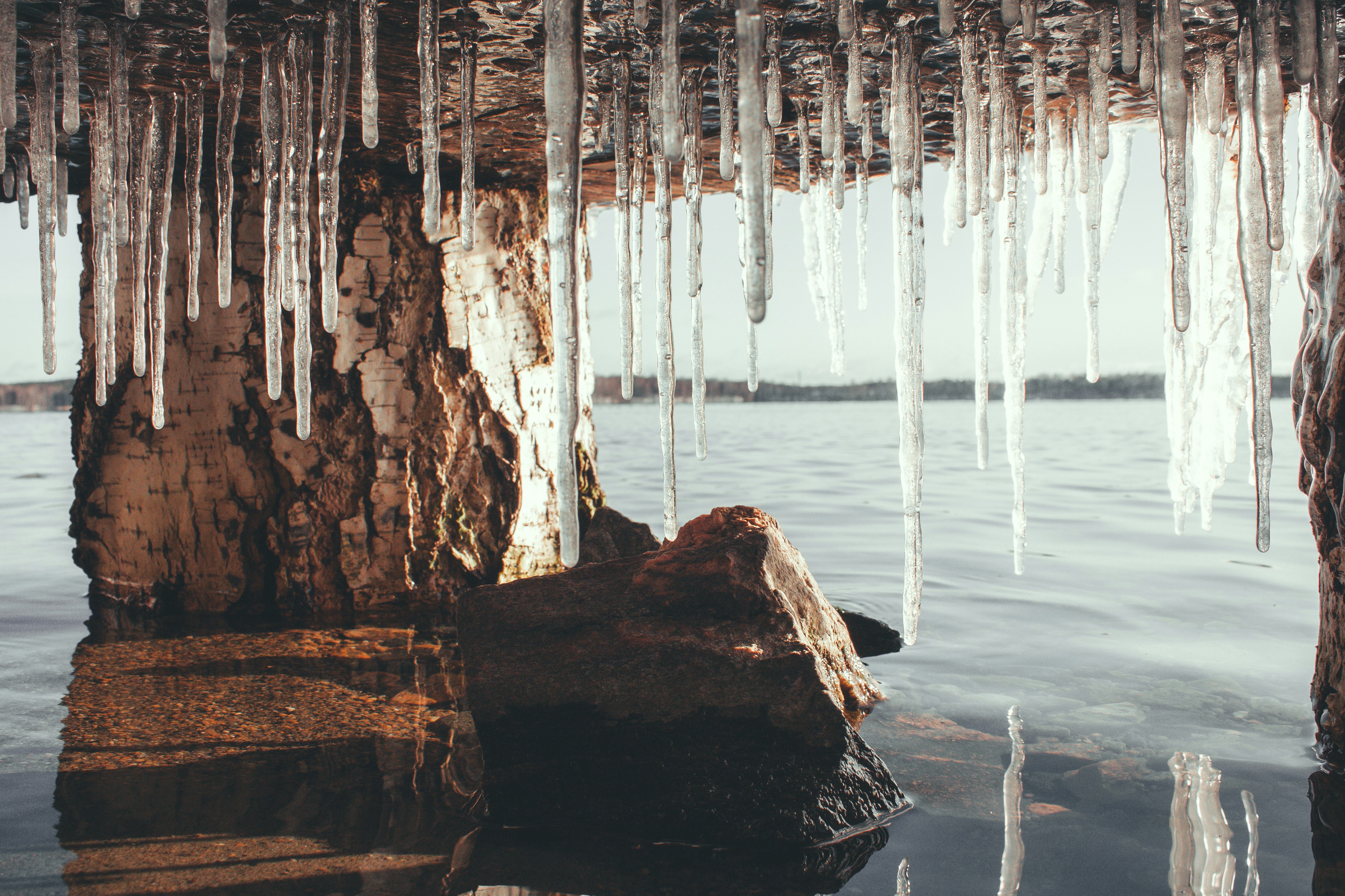 Icicles hang from a weathered wooden structure above a calm water surface, reflecting the serene landscape beyond.