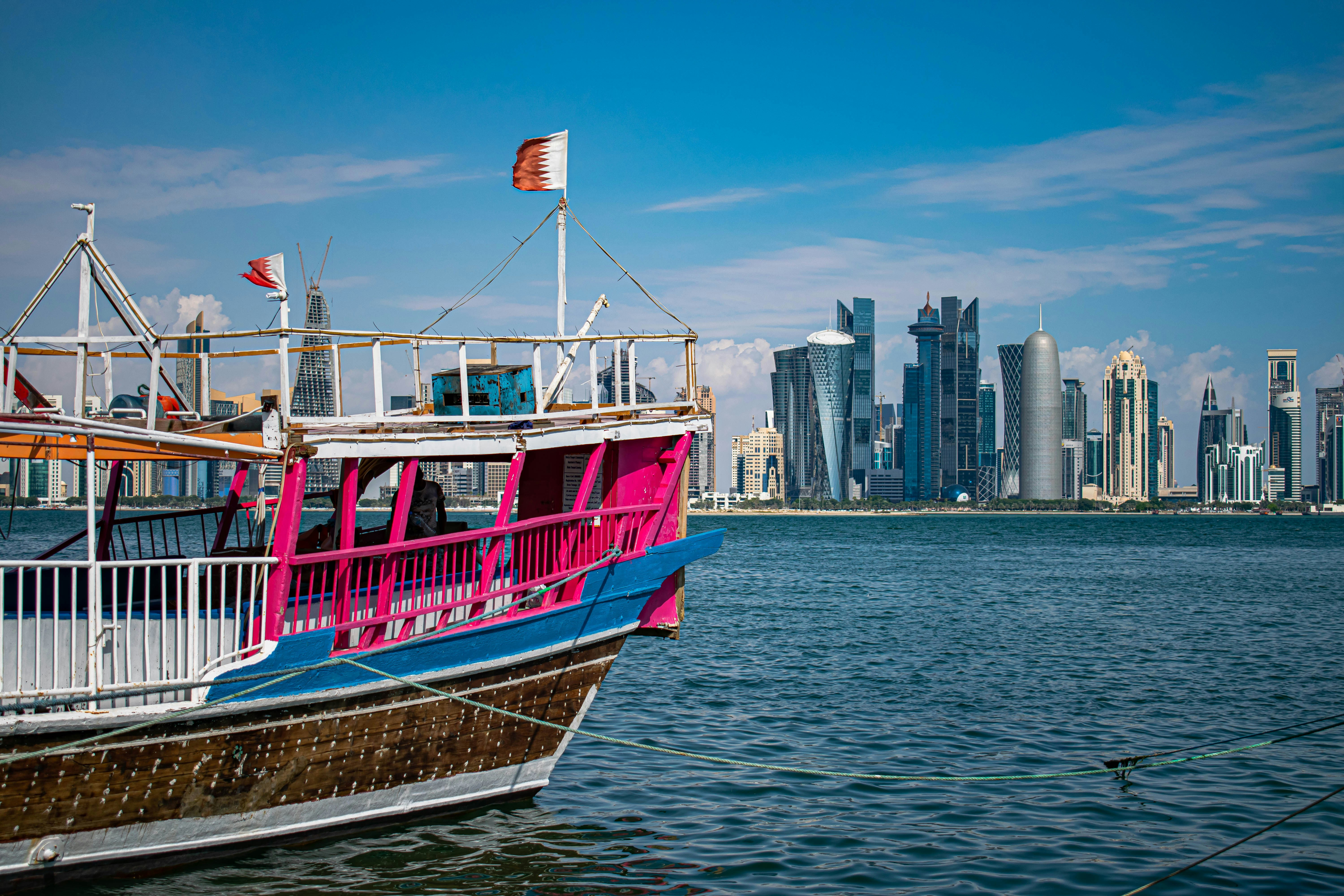 Traditional wooden boat with colorful accents on calm water with a modern city skyline in the background.