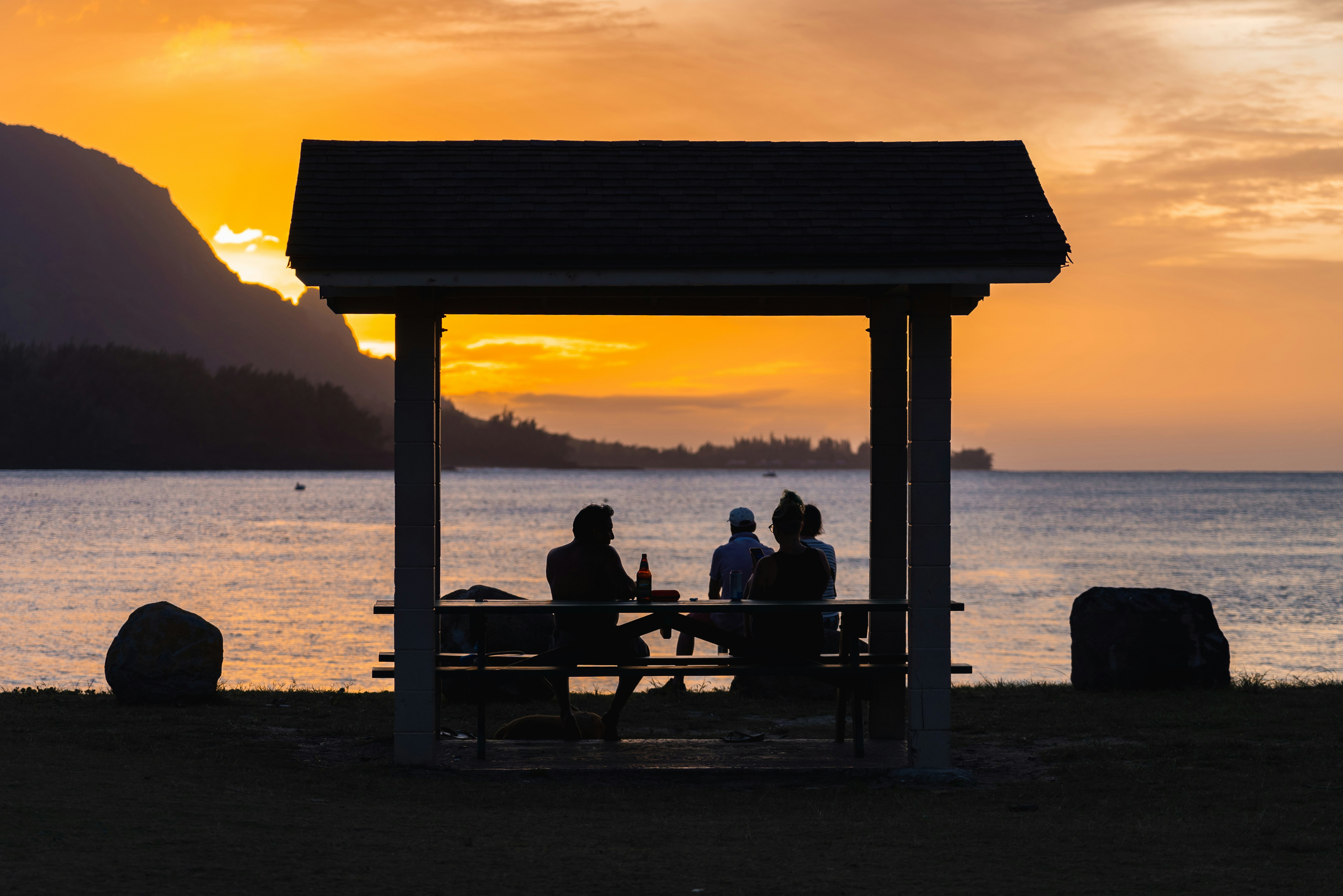 Silhouetted figures seated at a picnic table under a gazebo, watching a vibrant sunset over the ocean.