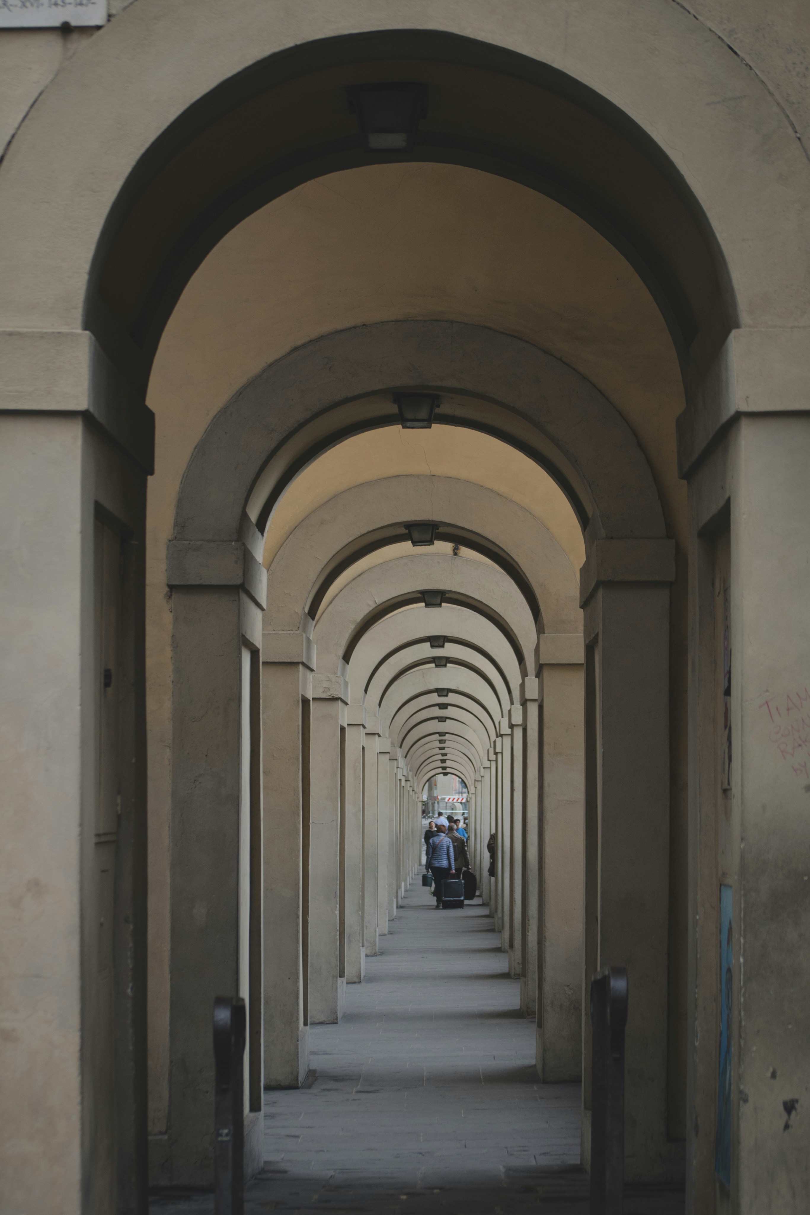 Archways leading down a corridor, with figures walking towards the end, creating a sense of depth and perspective.