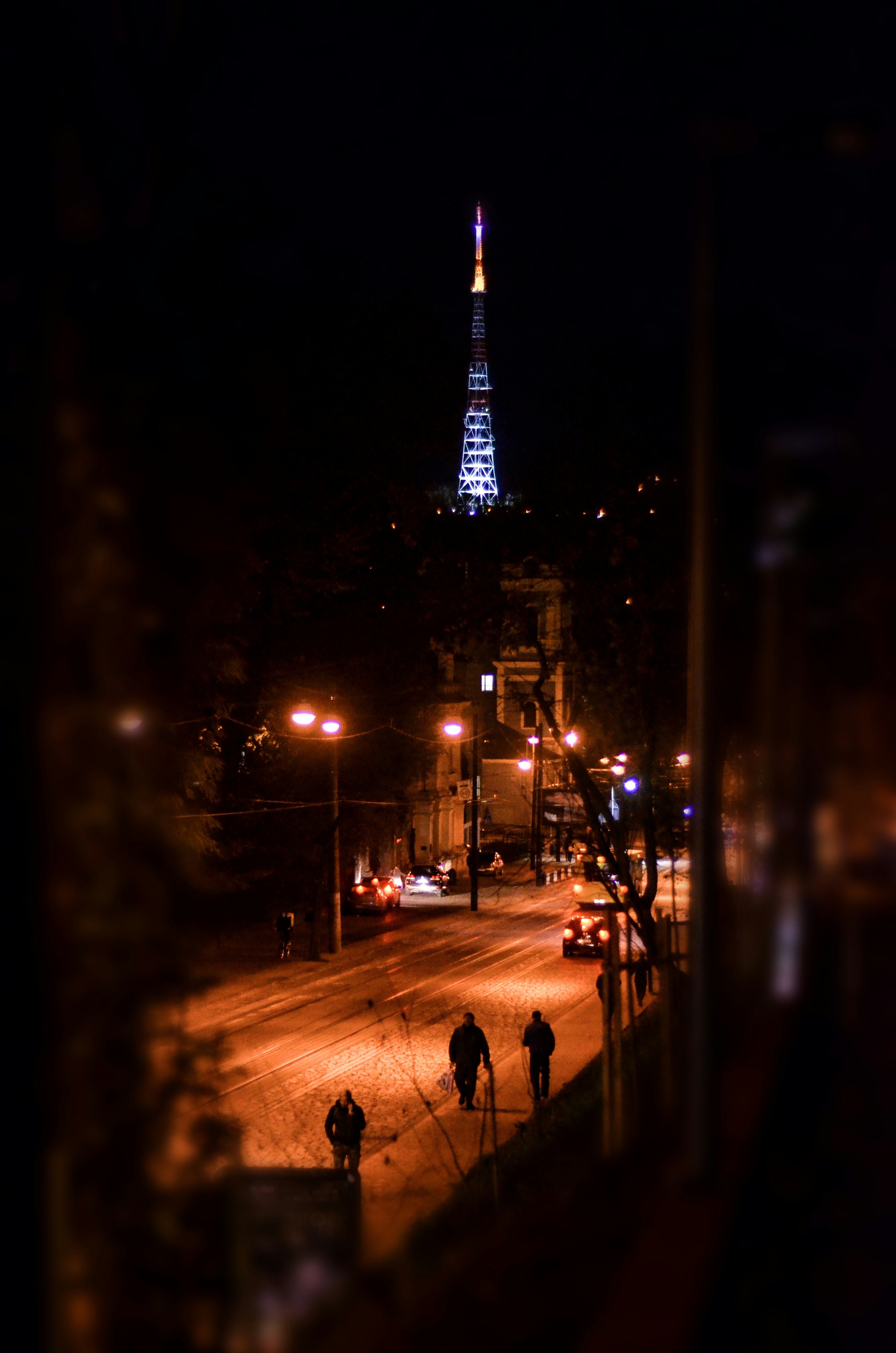 A vibrant city street under night sky, featuring a brightly lit tower in the background and pedestrians walking along the illuminated path.