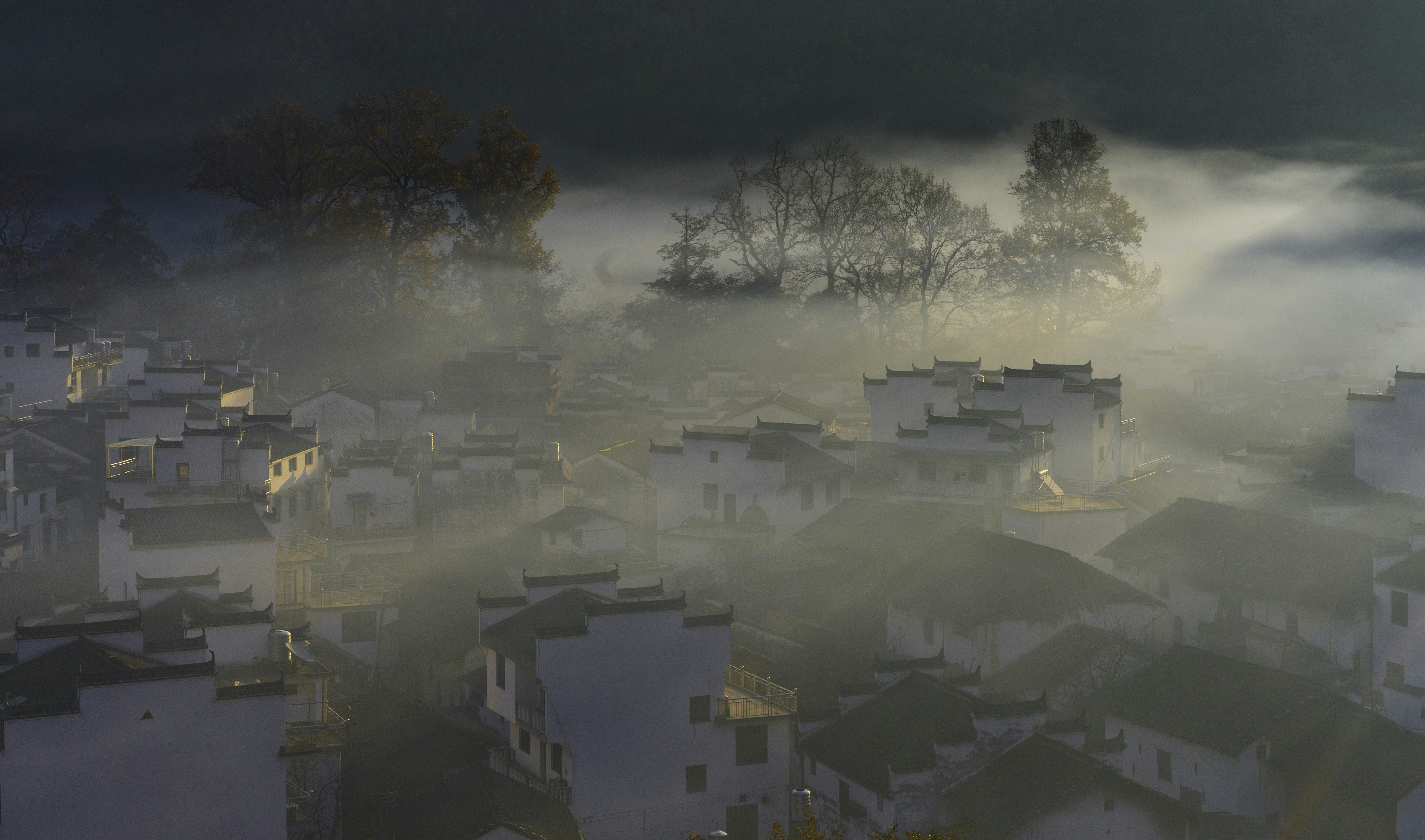 Mist swirls over a clustered hillside village of white-walled homes at dawn, with distant trees and sunlit fog. This atmospheric landscape photograph emphasizes quiet dawn light.
