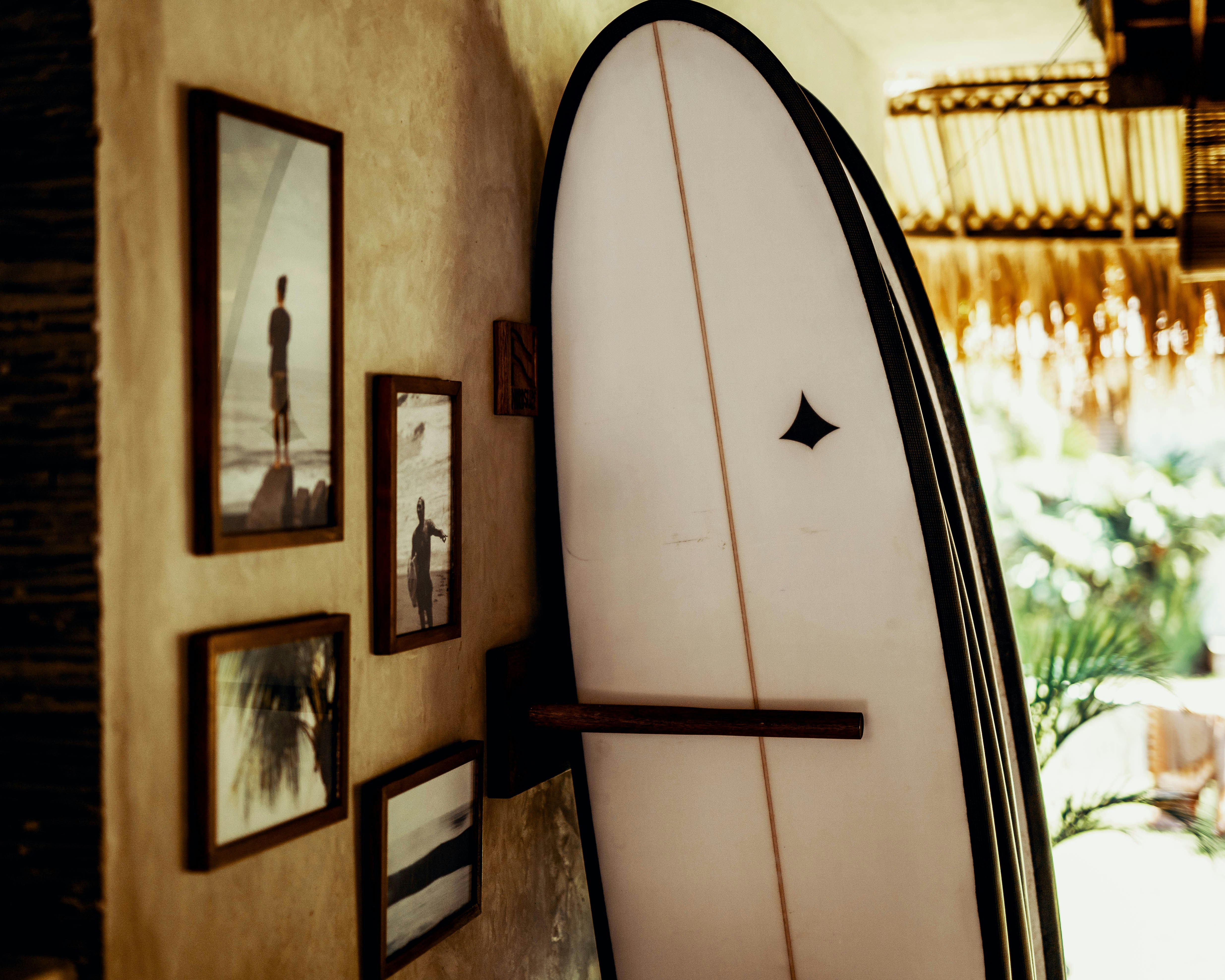 Surfboard mounted on a wall beside framed photographs, evoking a relaxed coastal atmosphere in a sunlit space.