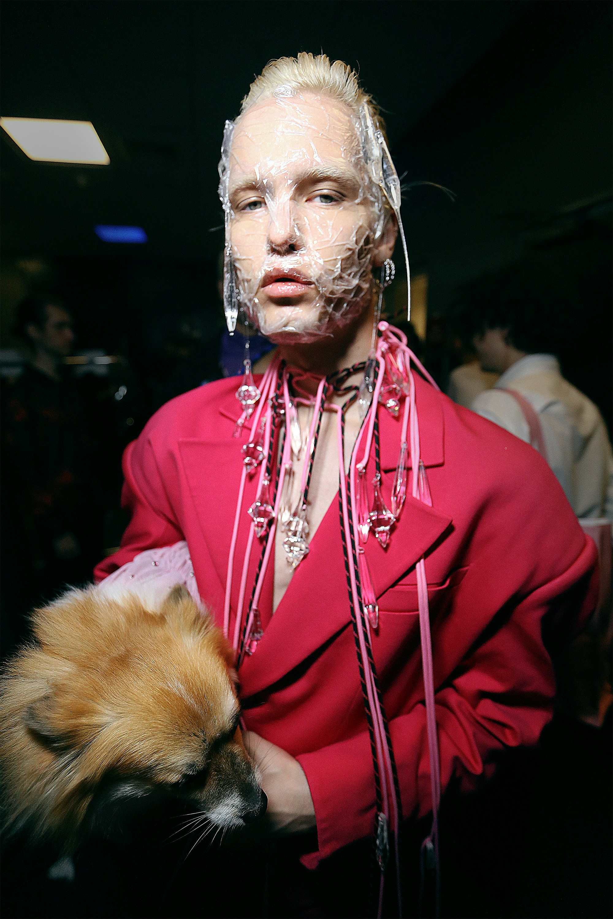 Model adorned in a striking pink ensemble with unconventional facial wrapping, cradles a small dog, creating a unique blend of high fashion and pet companionship.
