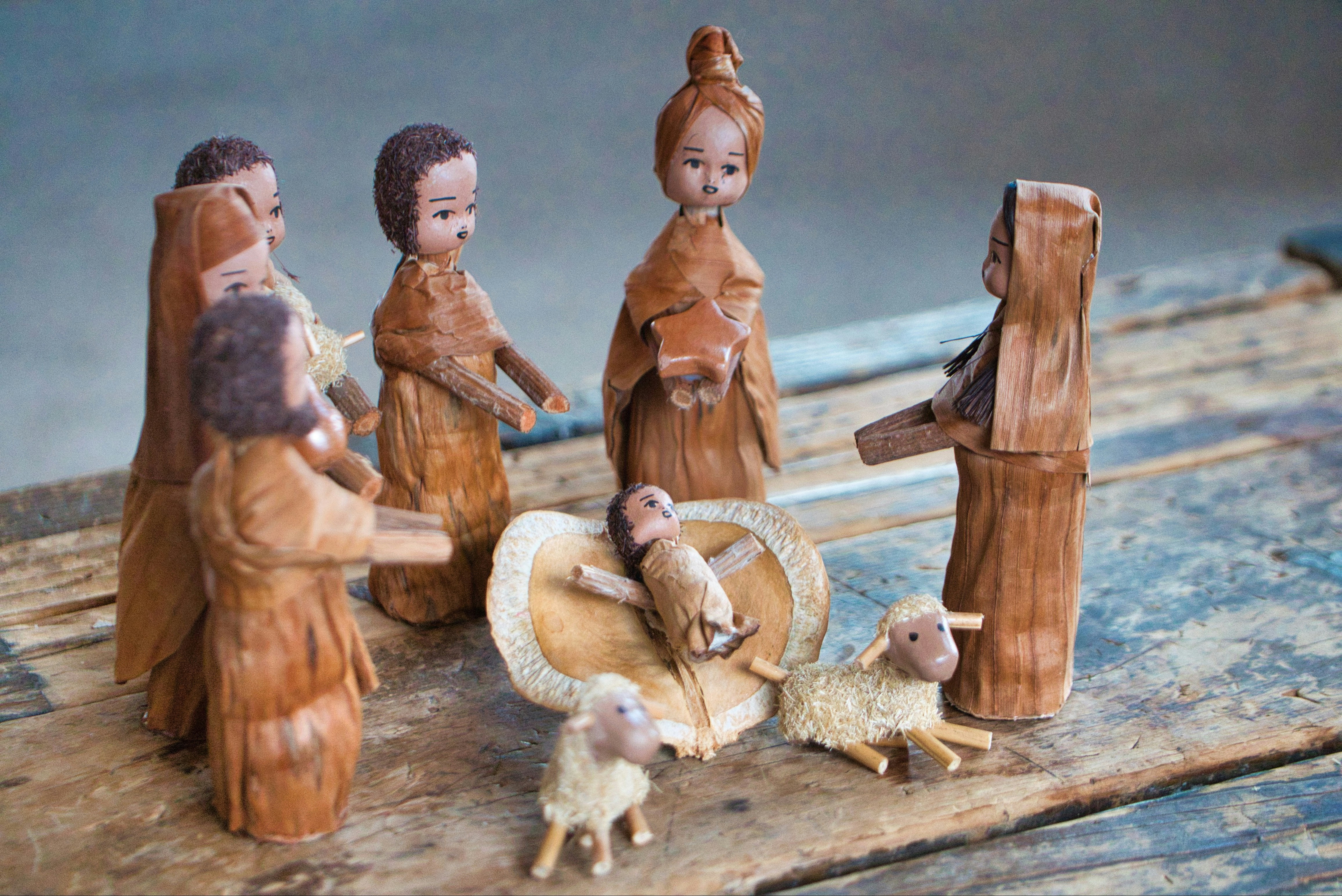 Handcrafted wooden figures depicting a nativity scene, featuring Mary, Joseph, and shepherds surrounding the infant Jesus in a cradle. The rustic setting enhances the artisanal craftsmanship.