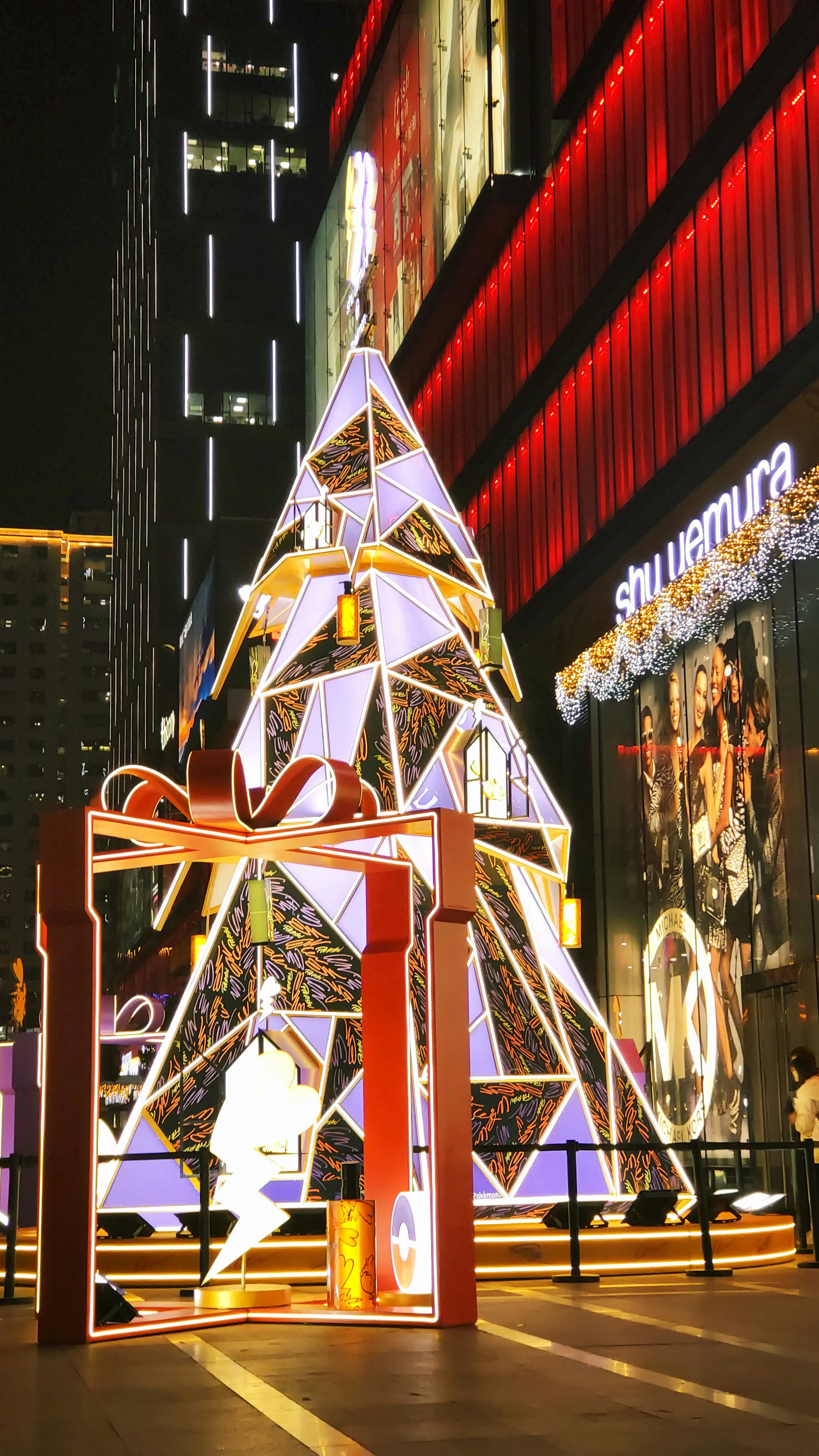 A vibrant holiday display featuring a geometric Christmas tree adorned with colorful lights and decorations, set against a backdrop of urban architecture.