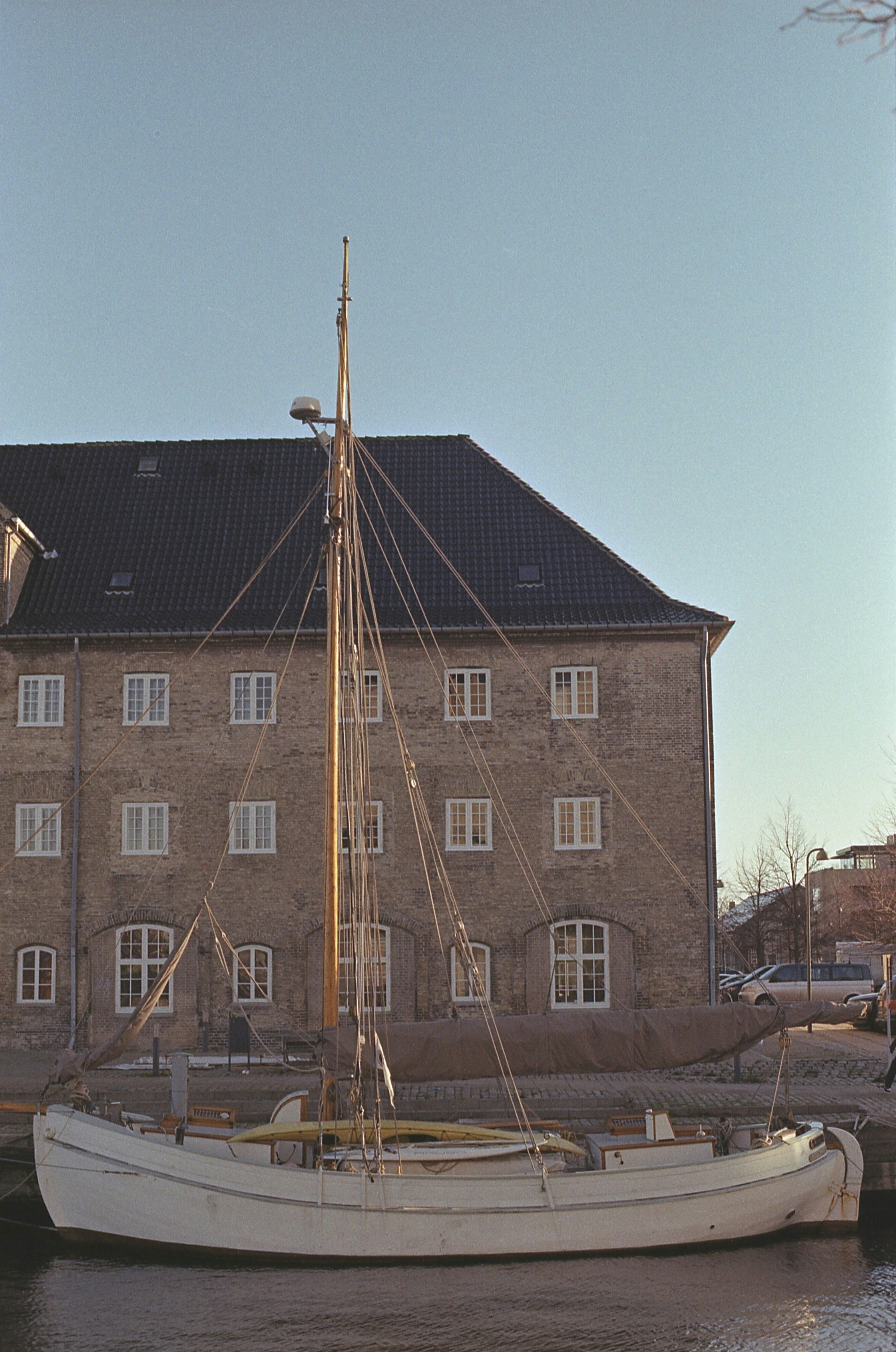 Sailboat docked along a brick waterfront with a tall mast and rigging, reflected in calm water.