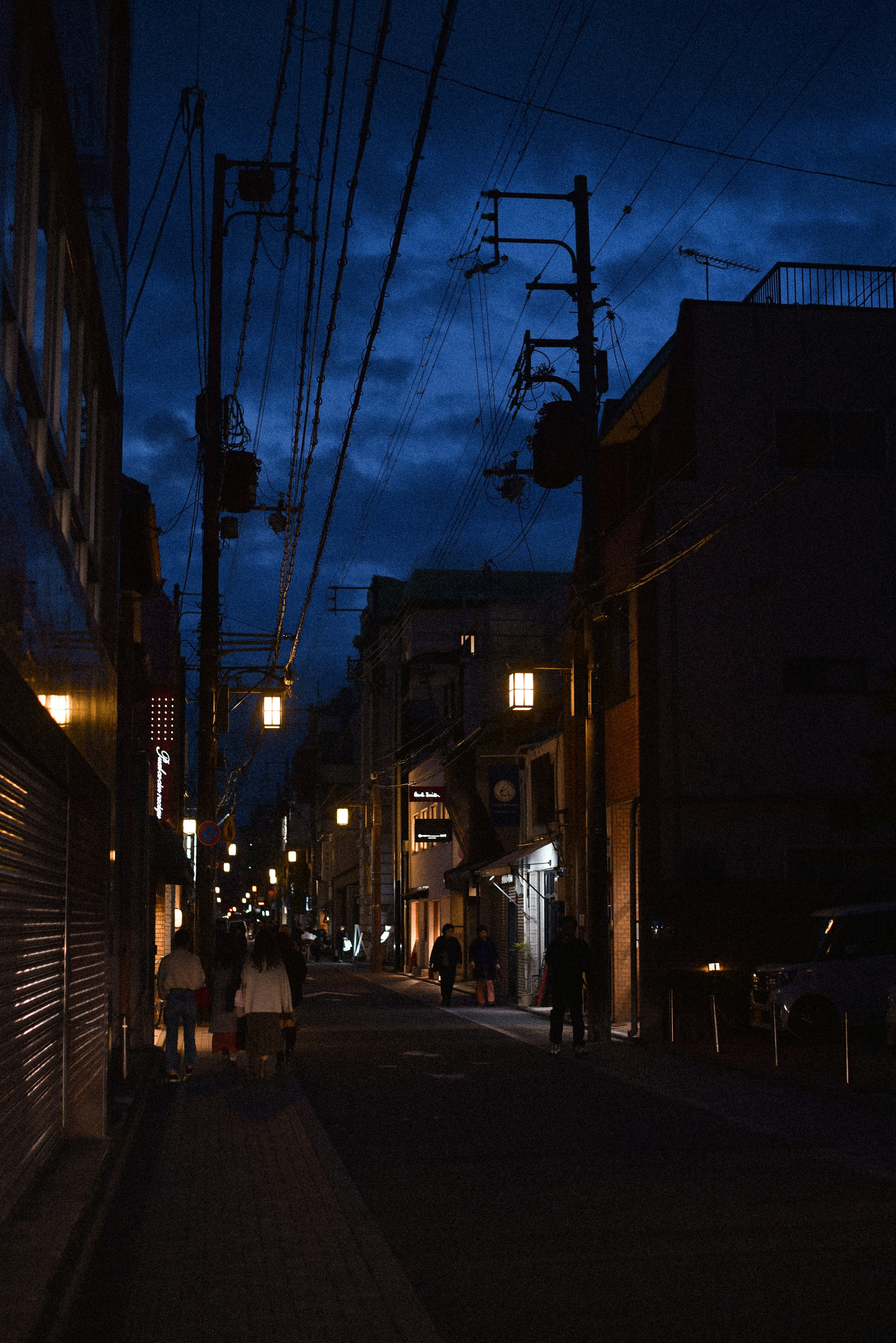 Dimly lit street scene at night, showcasing pedestrians walking under glowing lanterns amidst urban architecture. The atmosphere conveys a sense of calm and exploration.