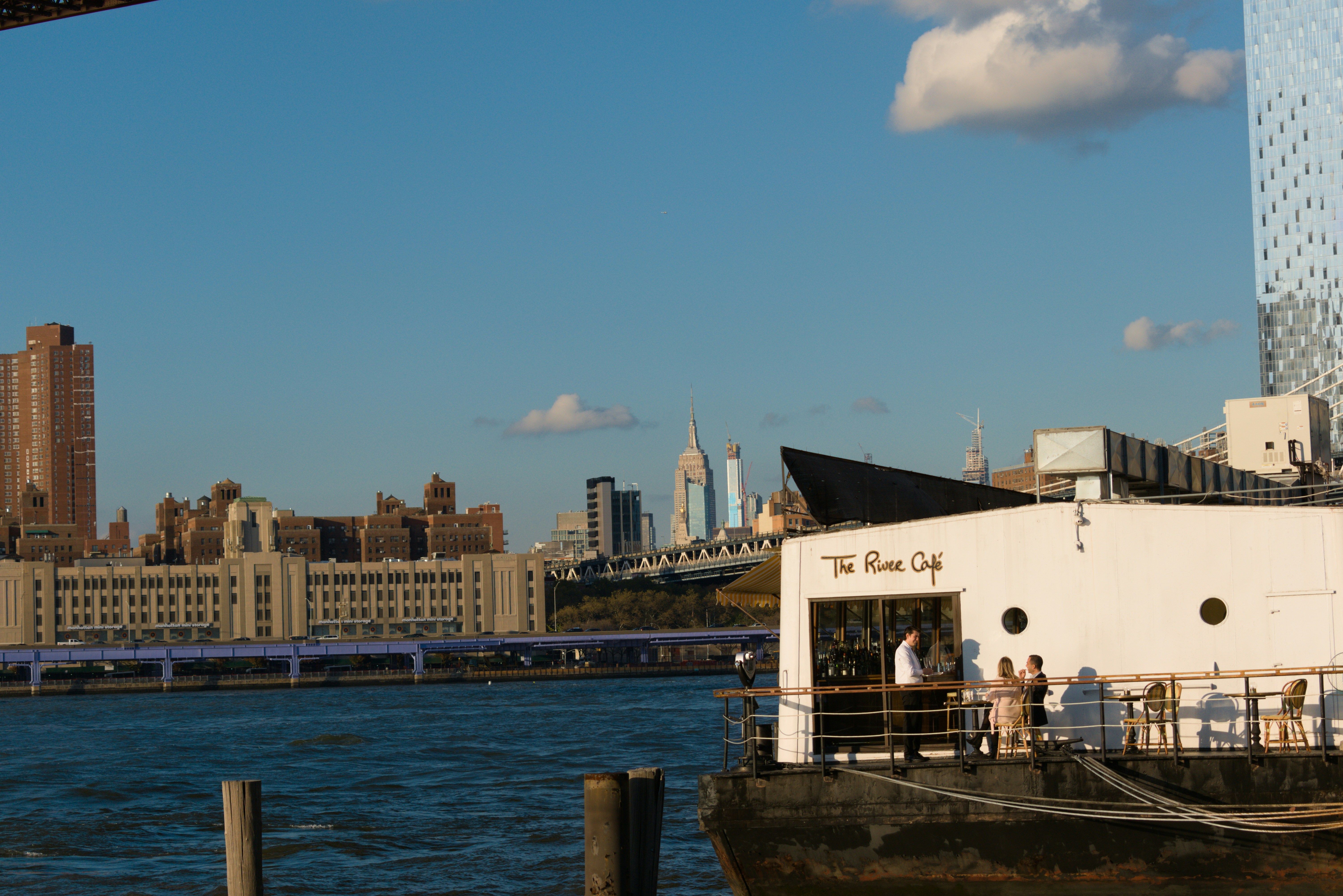 The River Café sits on the edge of the water, offering diners a scenic view of the Manhattan skyline, including iconic skyscrapers under a clear blue sky.