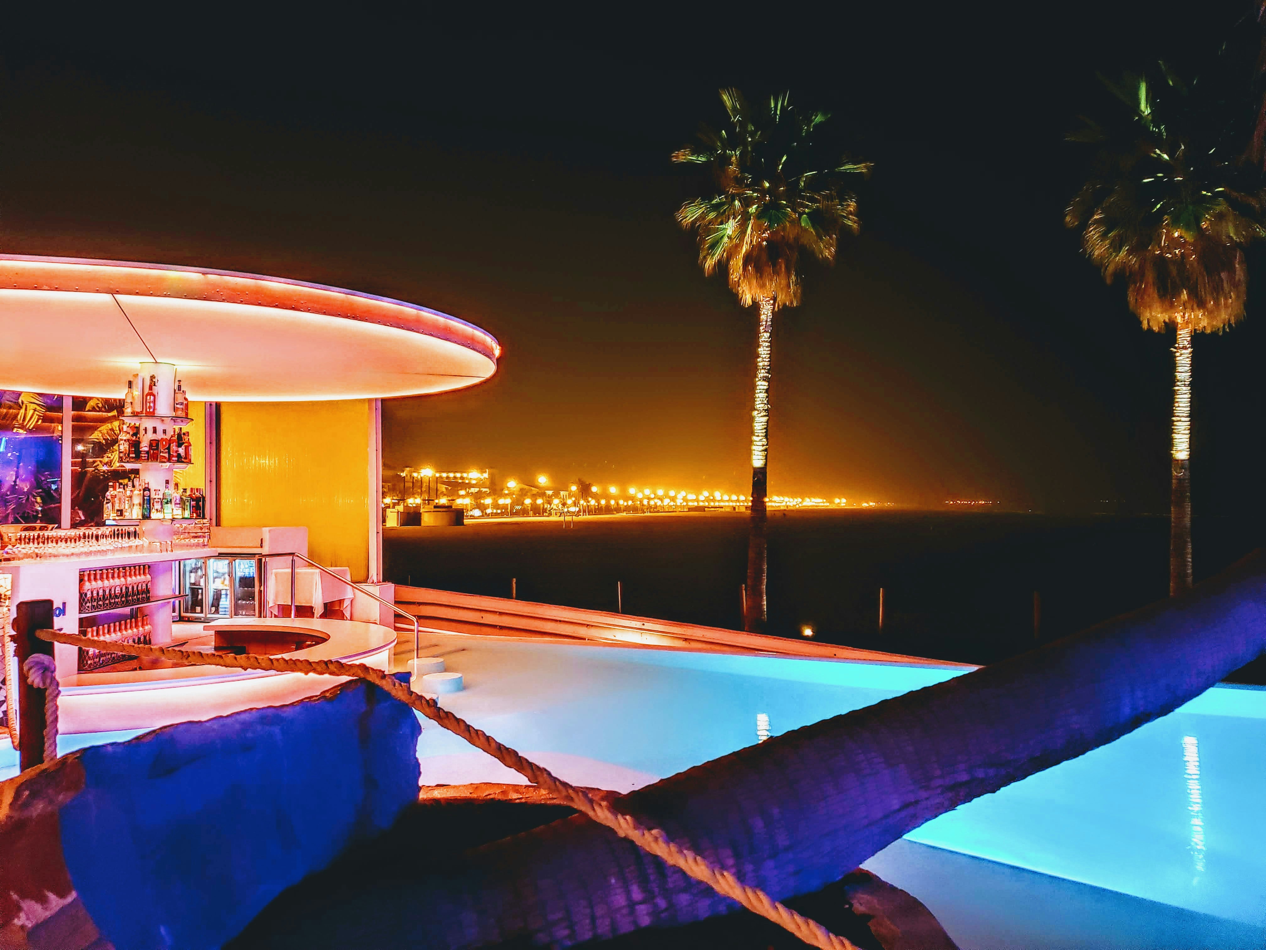 Illuminated poolside bar and palm trees at a seaside resort under night sky.