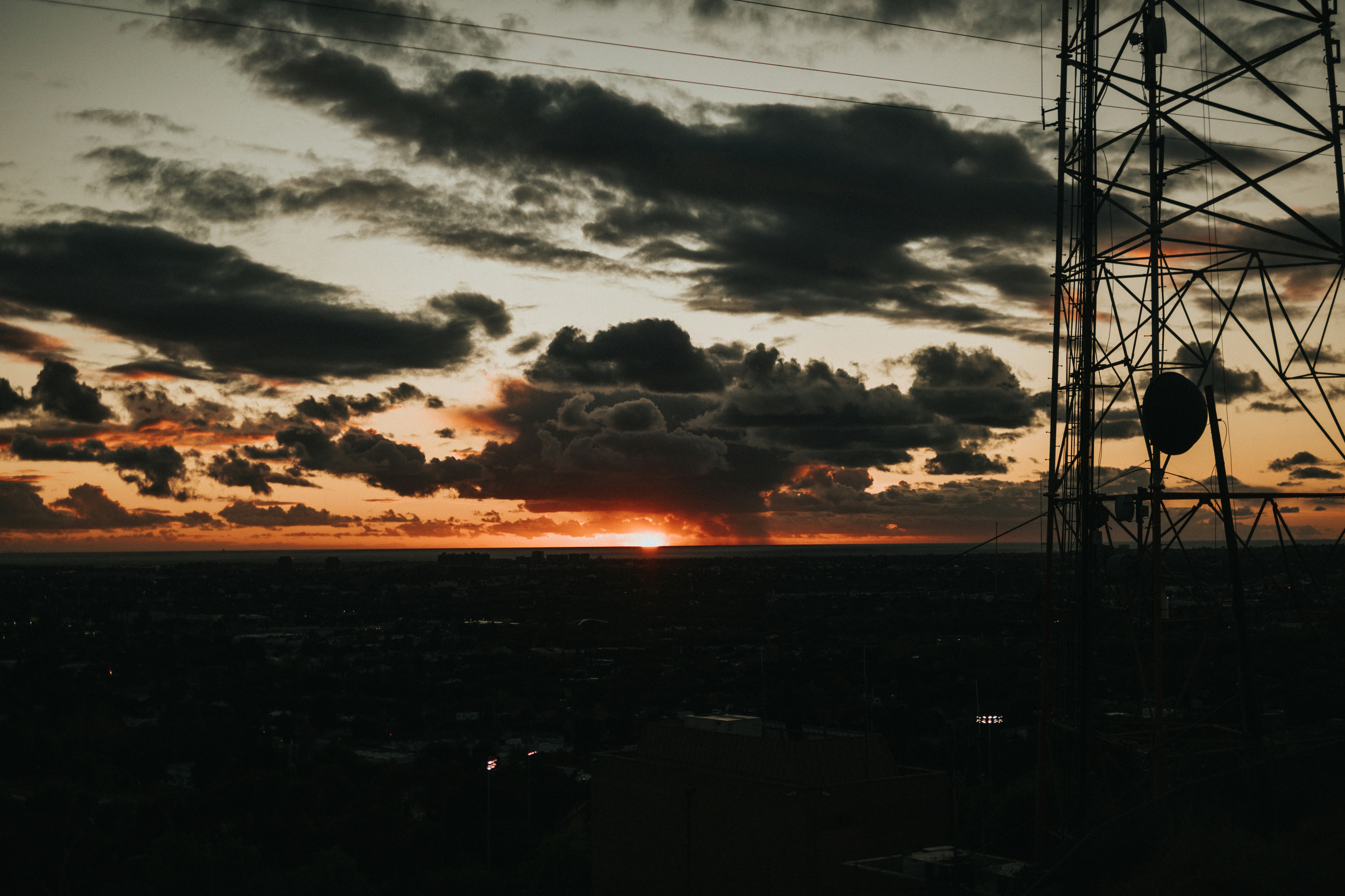 Silhouetted communication tower against a vibrant sunset, with clouds reflecting hues of orange and purple. The scene captures the transition from day to night.
