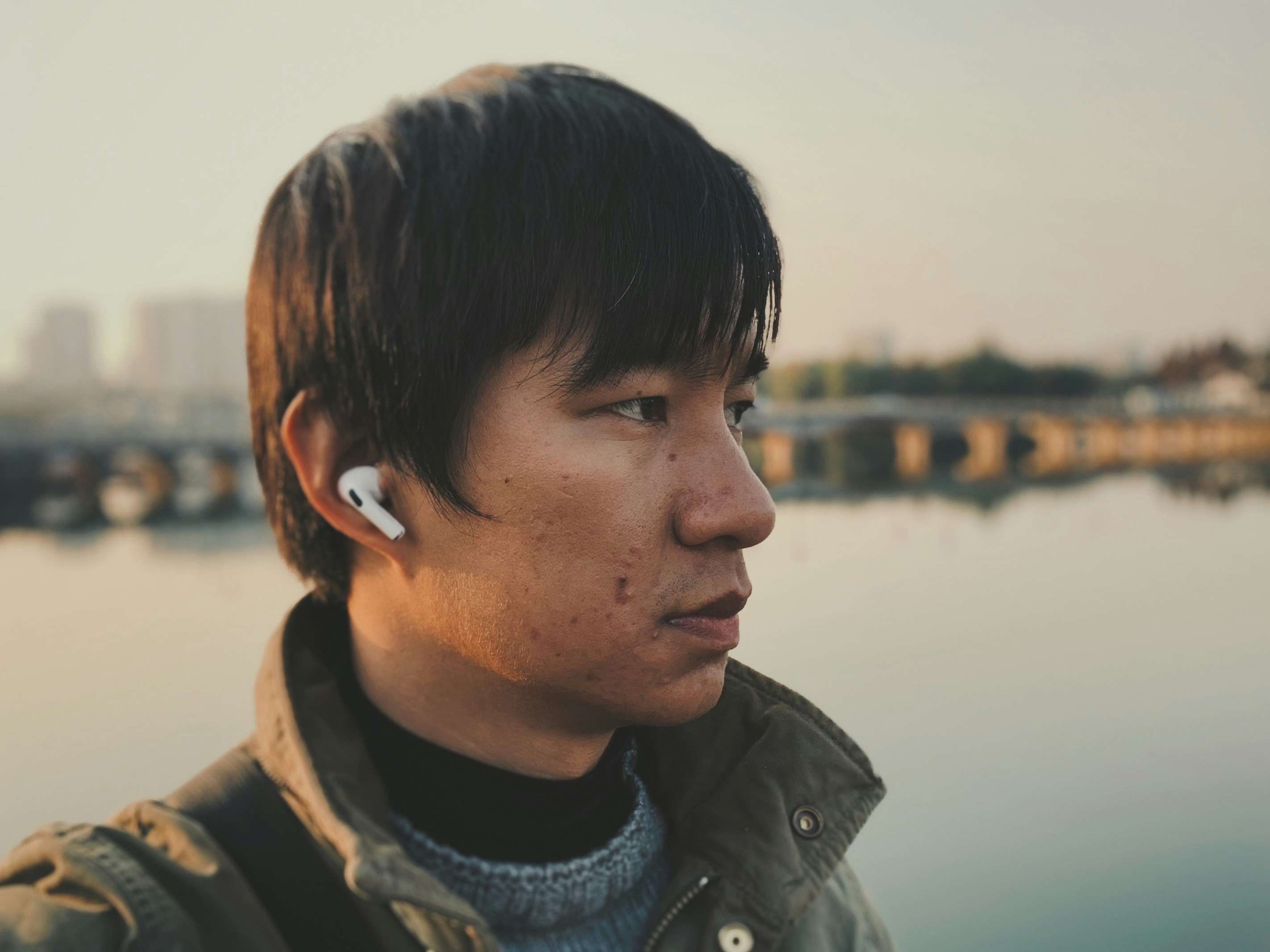 Profile of a young man wearing wireless earbuds, gazing thoughtfully towards a tranquil body of water at dawn.