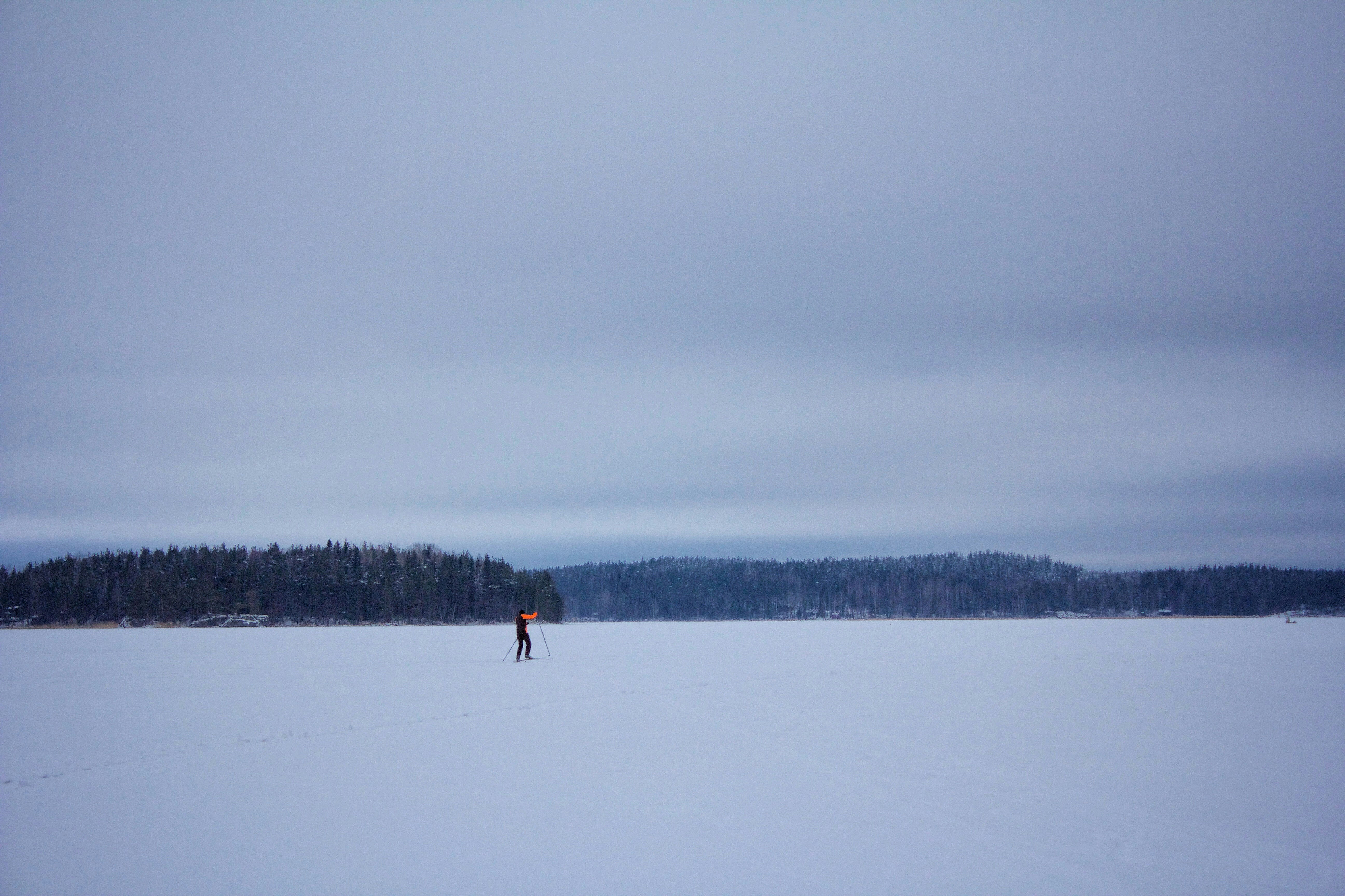 A lone figure skis across a vast, snow-covered lake under a muted sky, surrounded by a distant forest. The scene evokes a sense of tranquility and isolation.