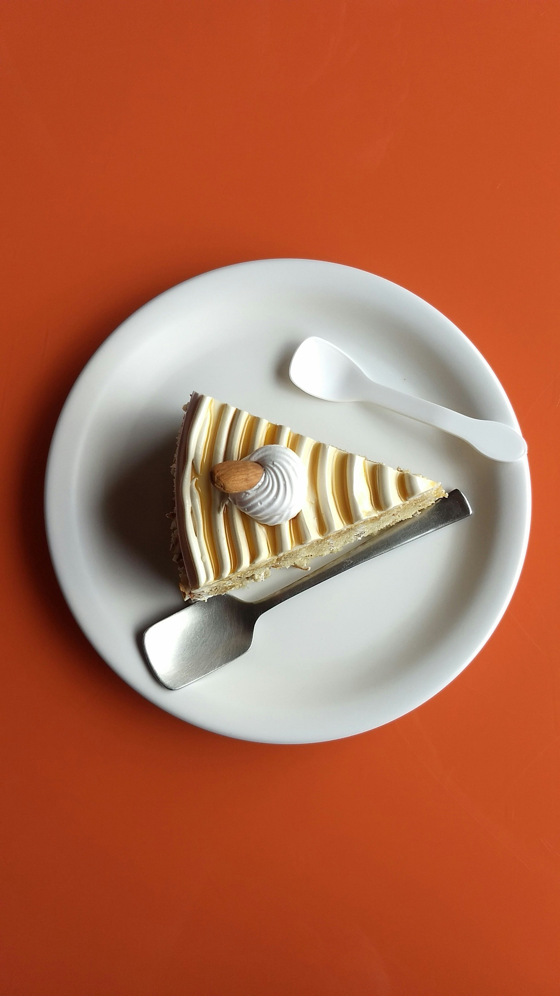 Creamy slice of cake topped with a swirl of whipped cream and almond on a white plate, accompanied by a stainless steel knife and a plastic spoon against a vibrant orange background.