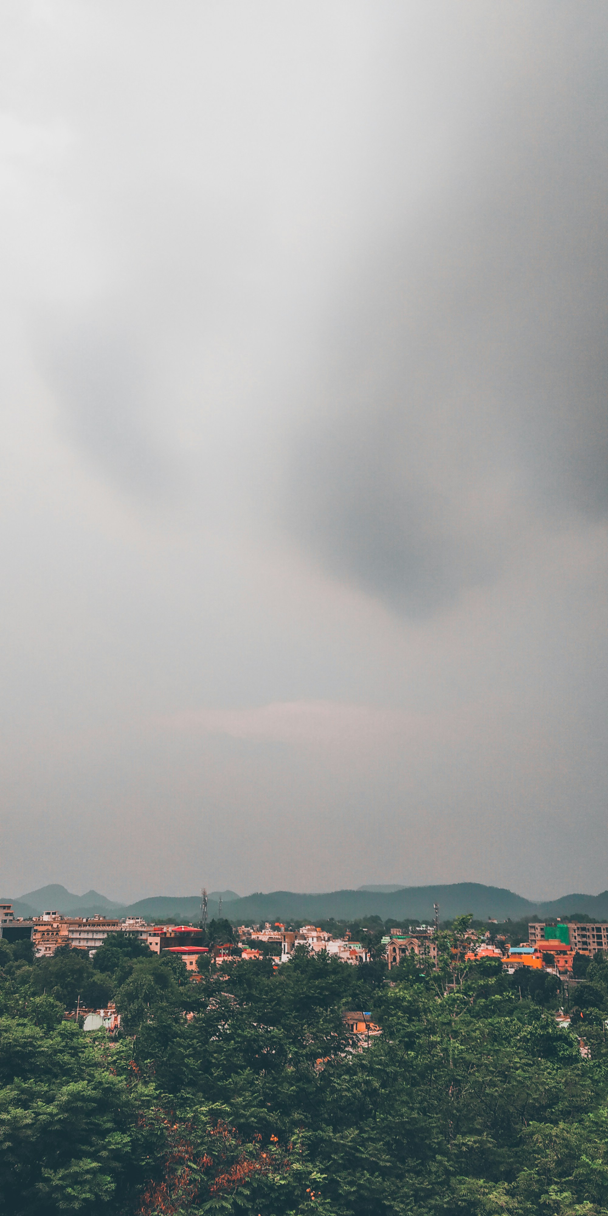 A panoramic view of a cityscape under a moody, overcast sky, framed by lush greenery and distant mountains.