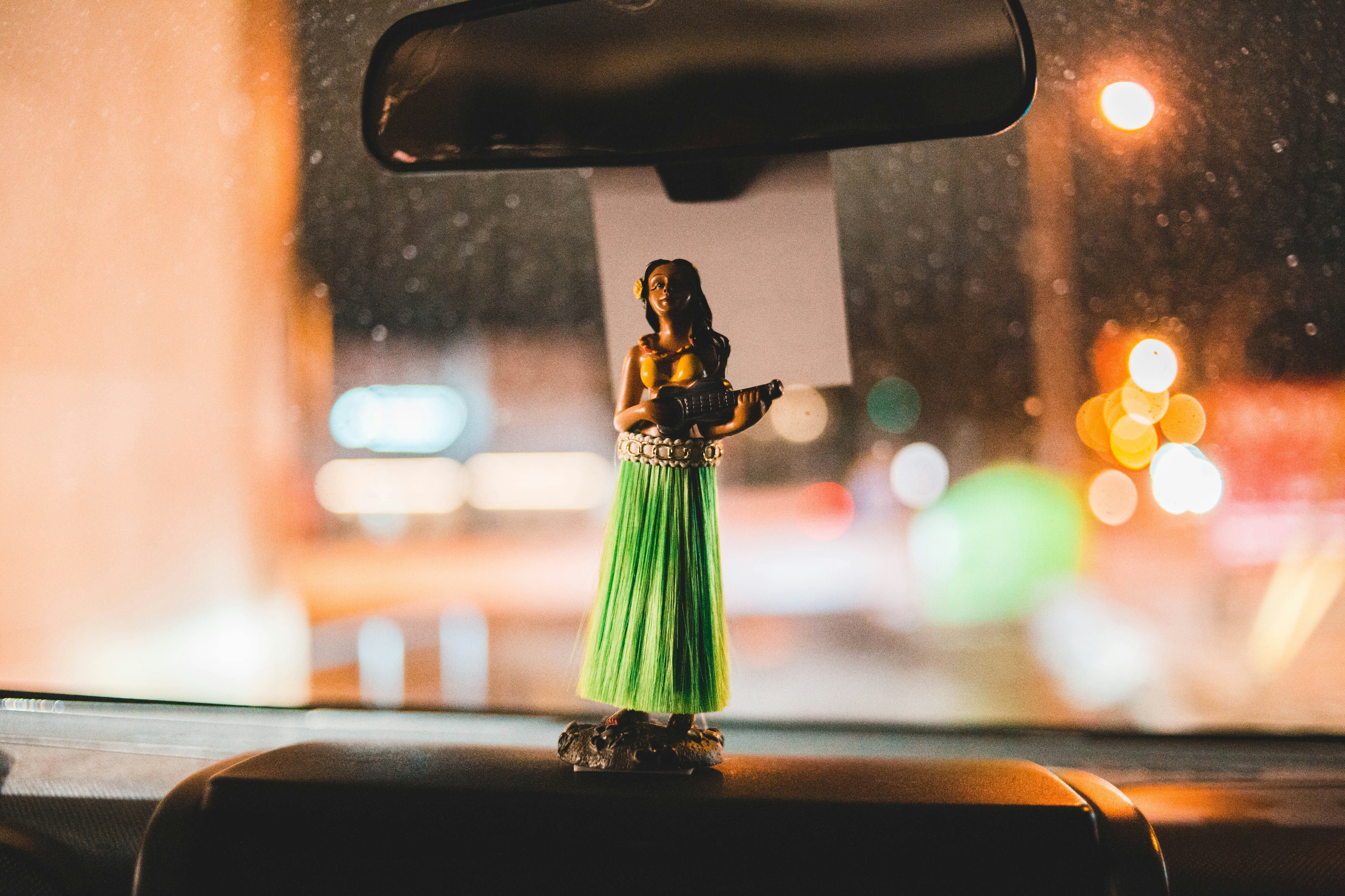 Tiny green dress, big city glow: a hood ornament scene on a rain-kissed windshield