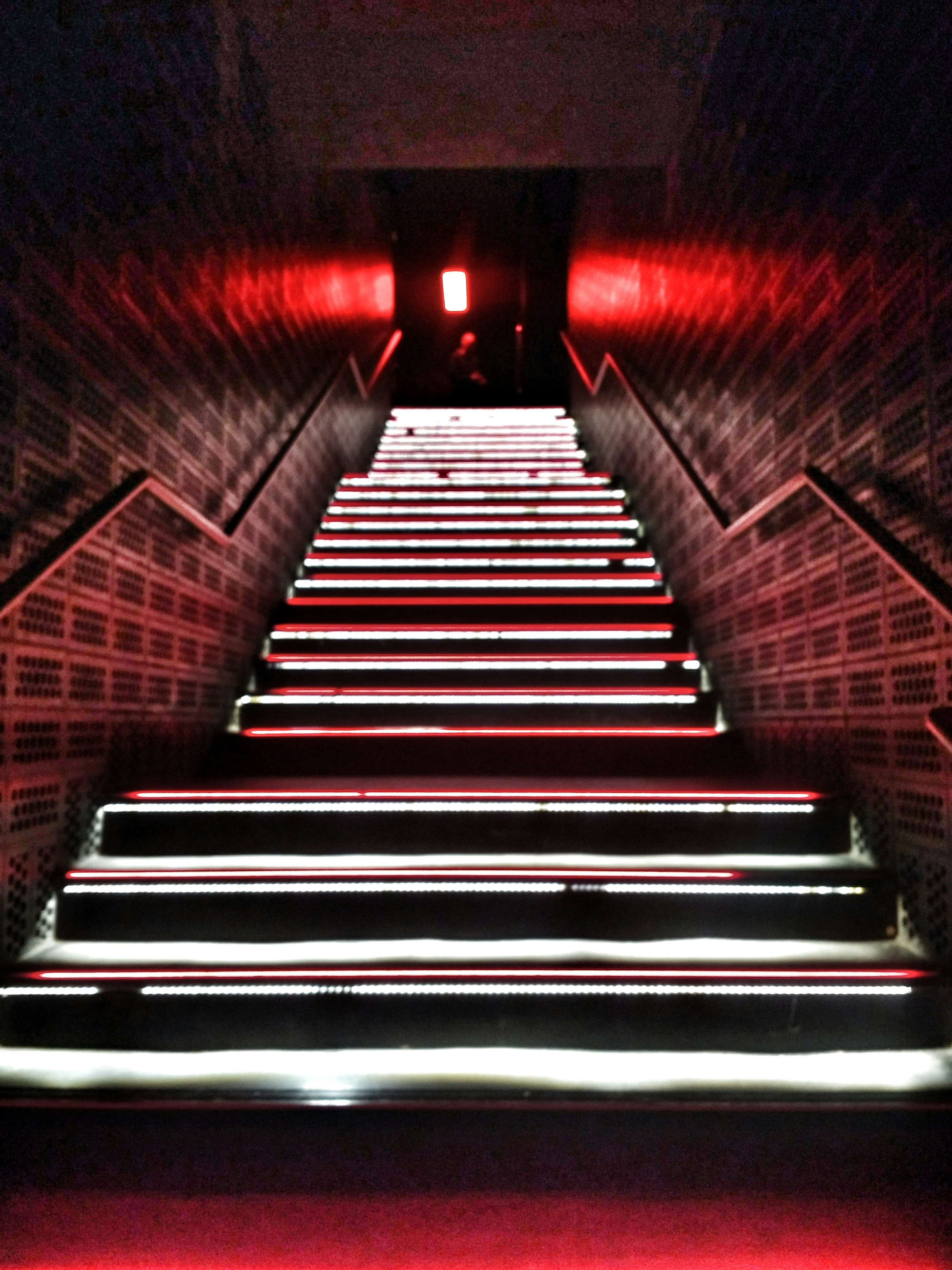 A staircase illuminated with red and white lights, leading upward into darkness, creating a dramatic and atmospheric effect.