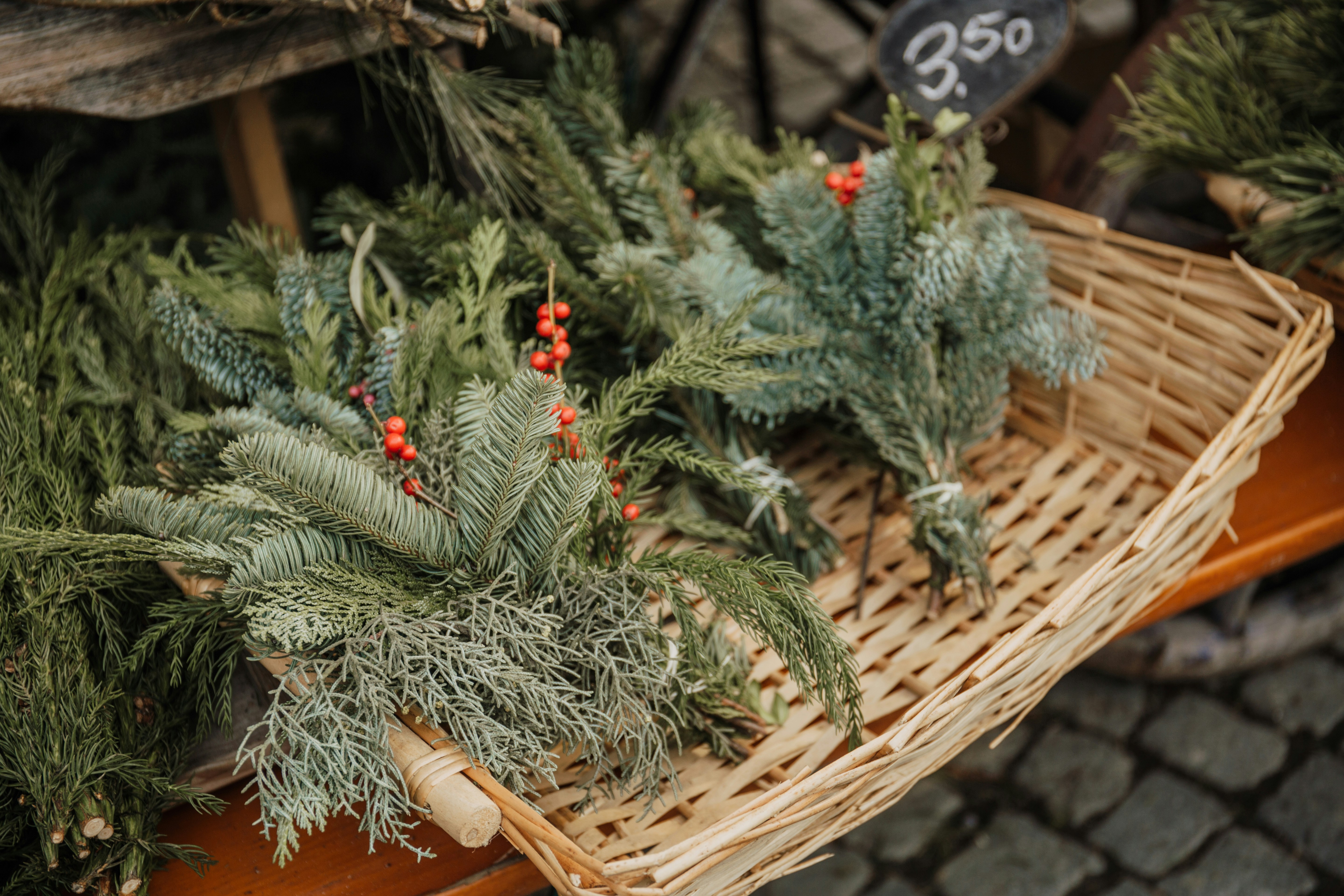 Wicker basket on a cobblestone surface holds blue-green evergreen sprigs with red berries, creating a textured holiday display.