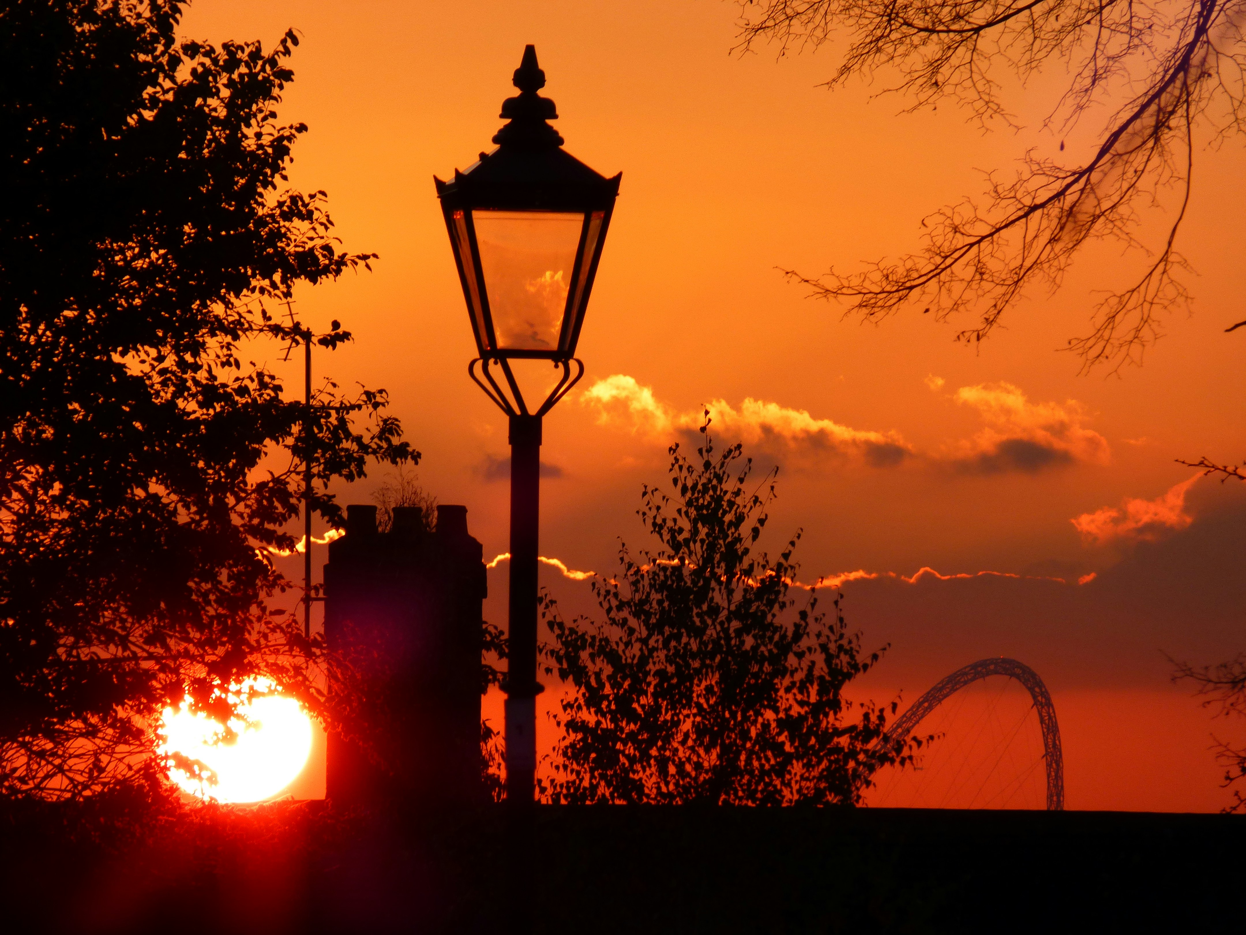 Lamppost silhouetted against a vibrant sunset sky with trees and distant structures.