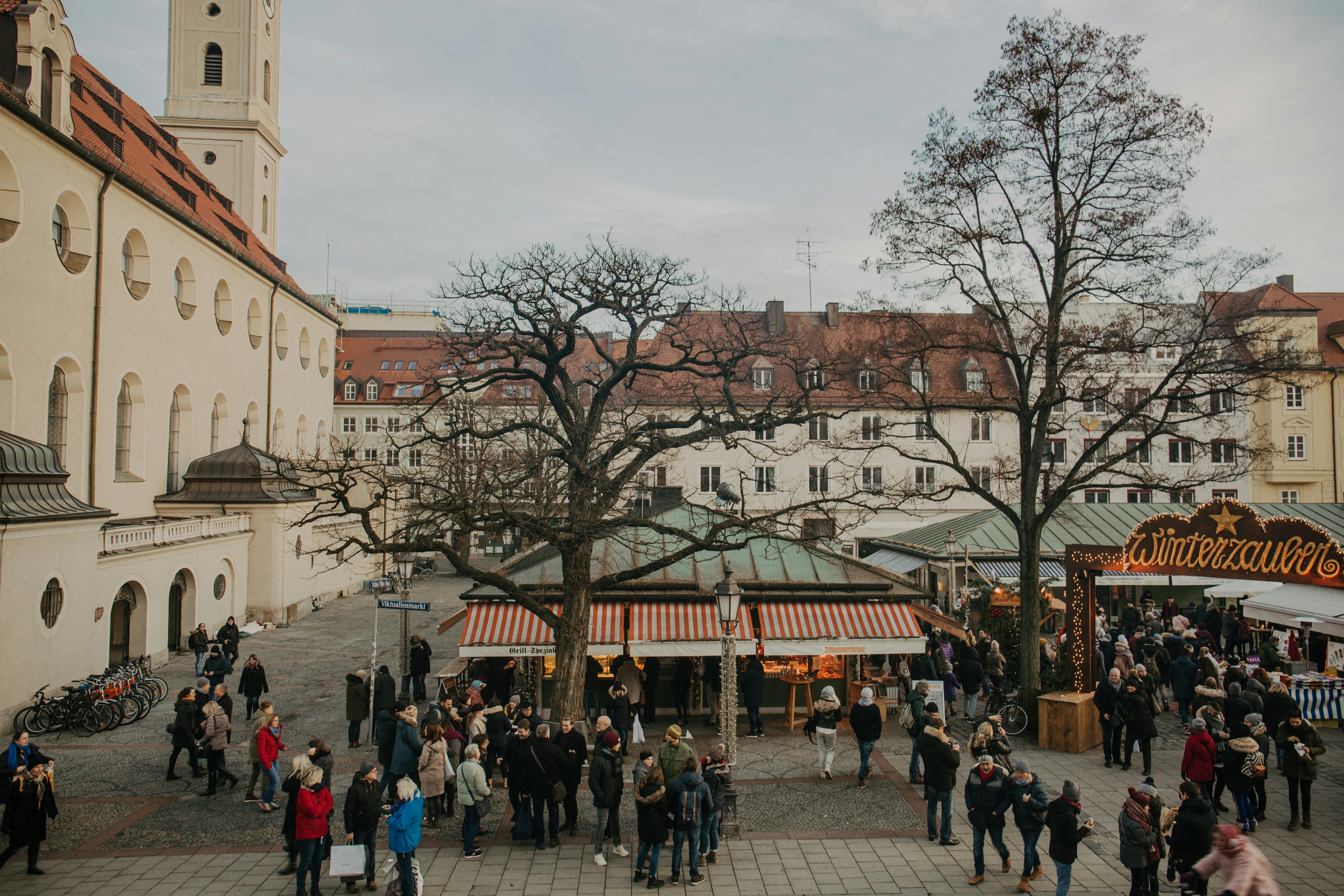 Crowd gathered at a winter market featuring festive stalls and a central tree, surrounded by historic architecture. The atmosphere is lively and inviting.
