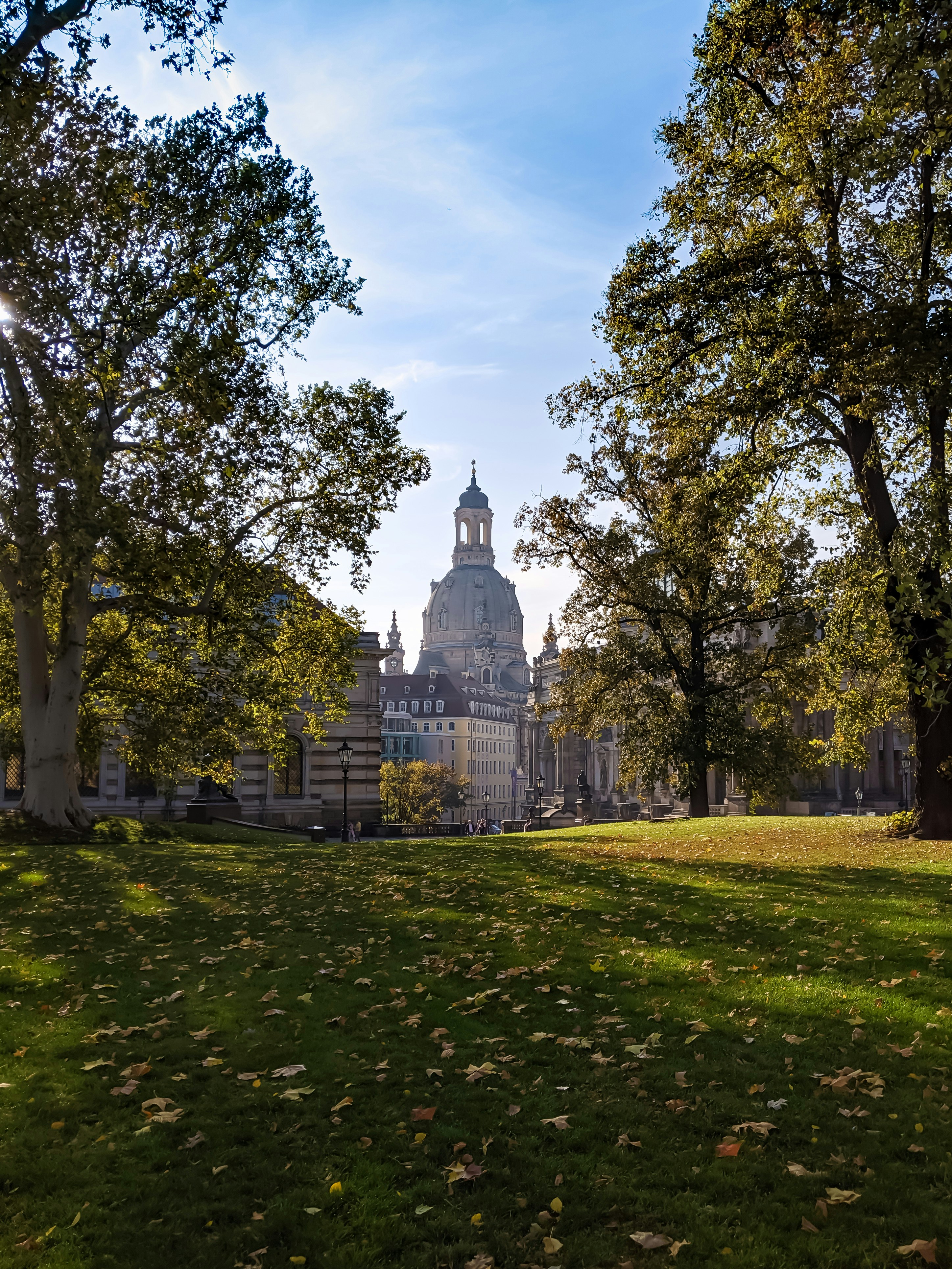Historic building framed by lush trees in a sunlit park, showcasing autumn leaves scattered on the ground.