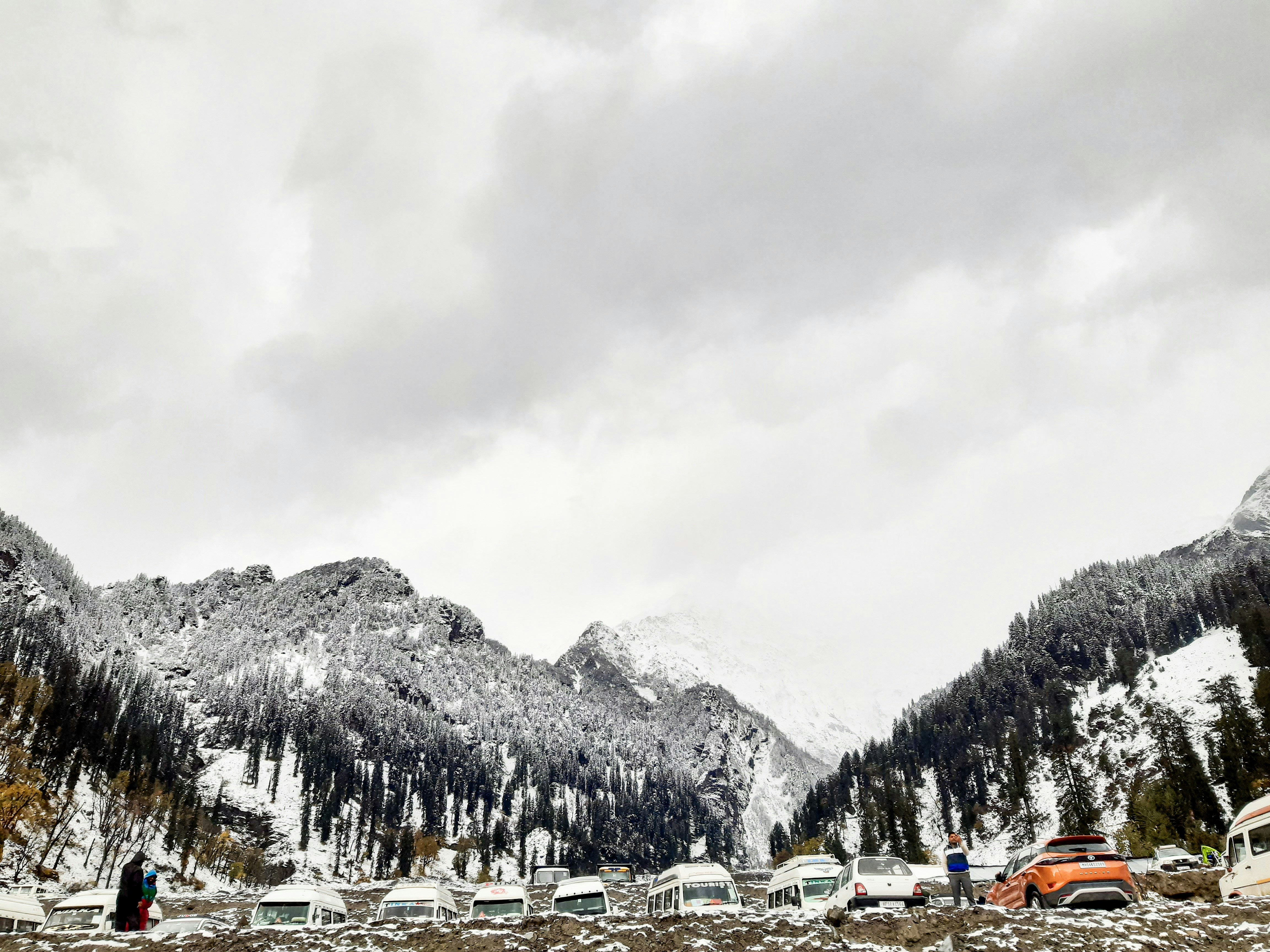 Snow-covered mountains loom over a parking area filled with vehicles, hinting at a winter adventure. A lone figure walks amidst the serene landscape.
