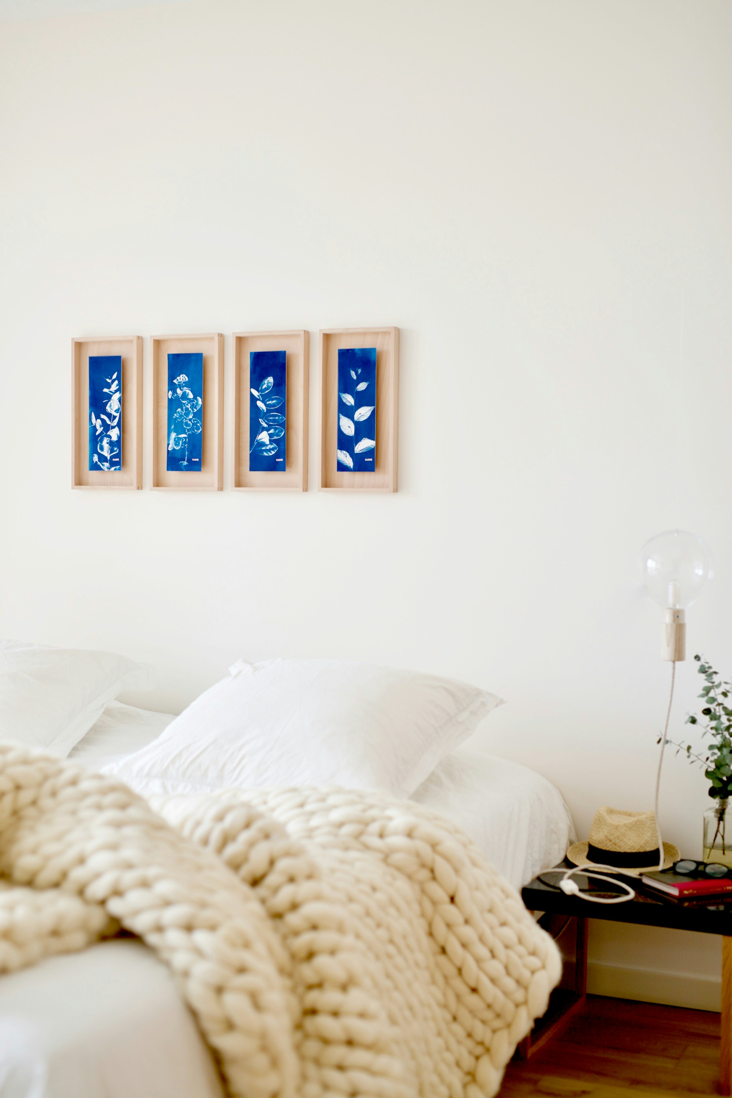 Photograph of four blue botanical prints in wooden frames arranged on a white wall above a cozy bed with a chunky knit blanket.