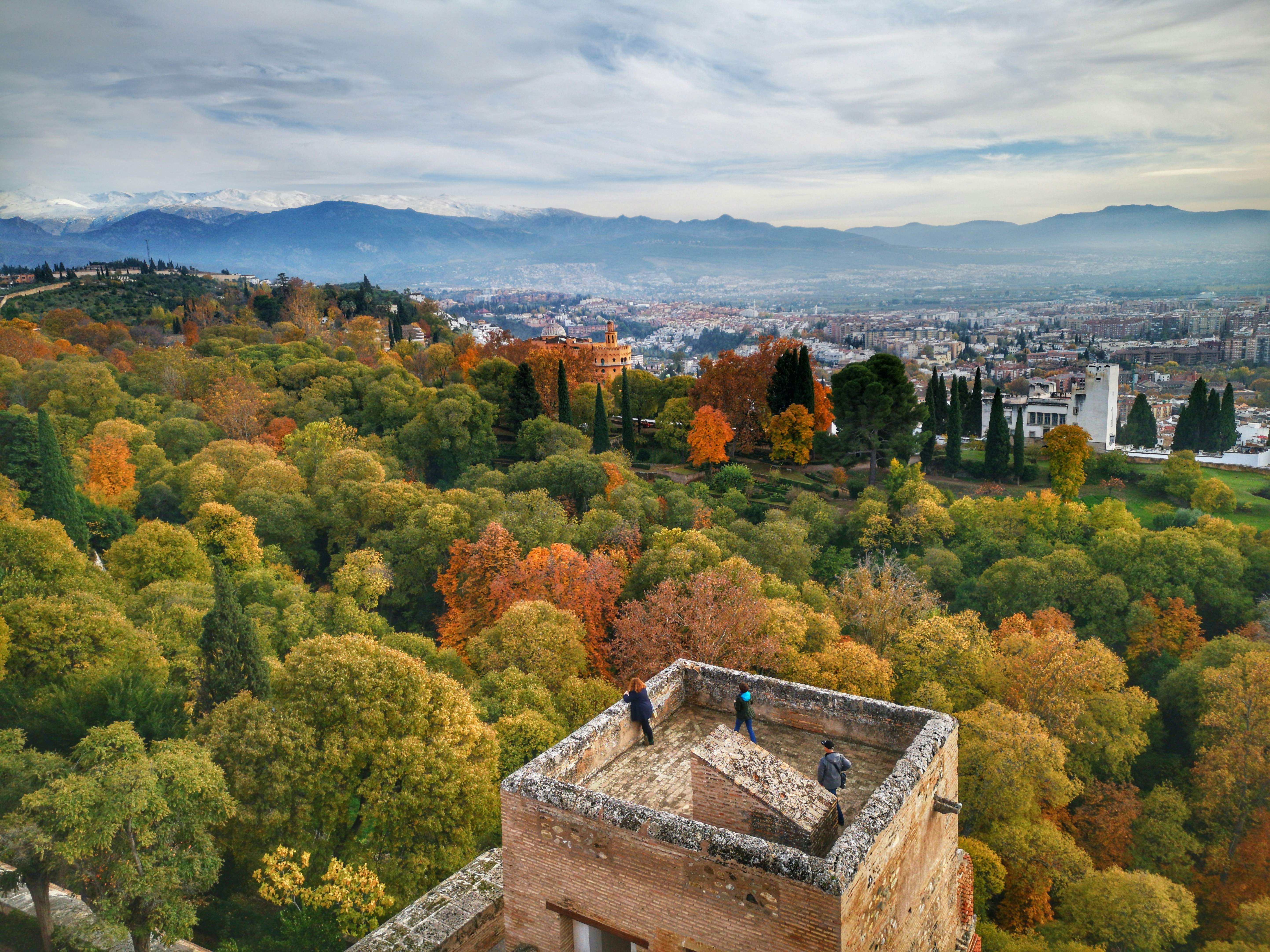 Vibrant autumn foliage surrounds the Alhambra with distant mountains under a cloudy sky.