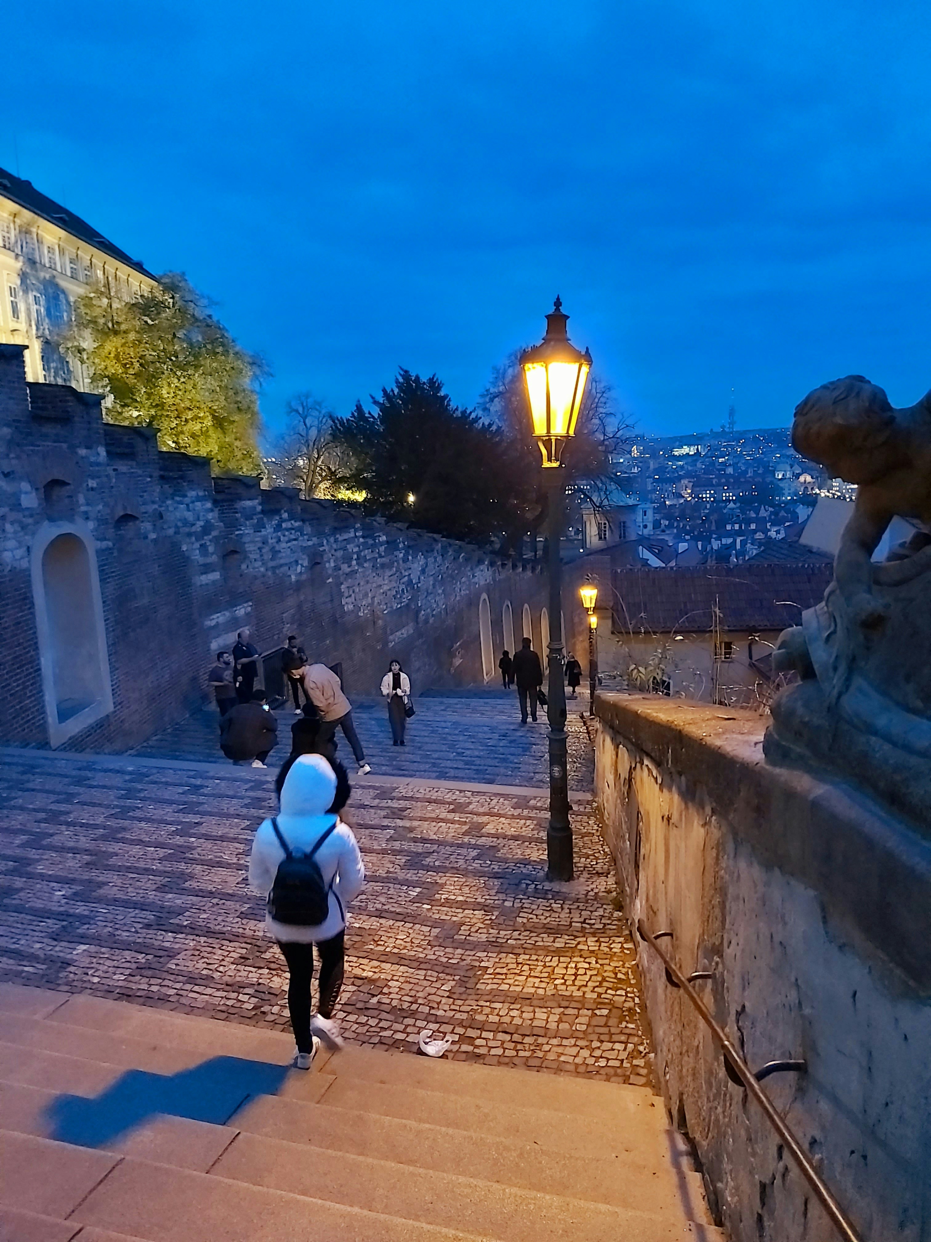 A blue-hour photograph of a cobblestone stairway lined with warm lamps, with a distant city glow in the background. A person in a white hoodie descends while bystanders linger along the wall.