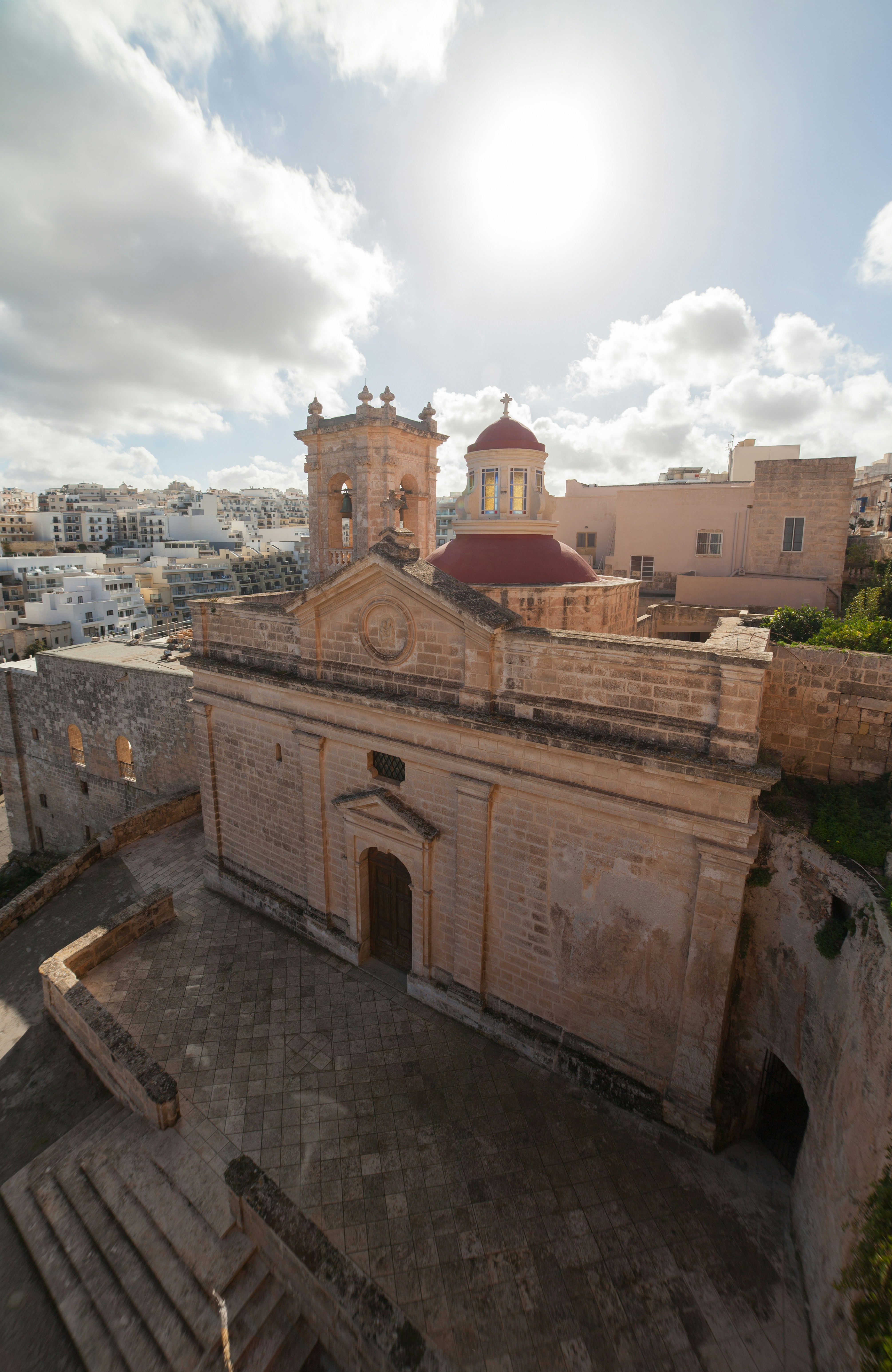 Historic church with a red dome and bell tower, surrounded by modern buildings under a bright sky. Stone steps lead to the entrance.