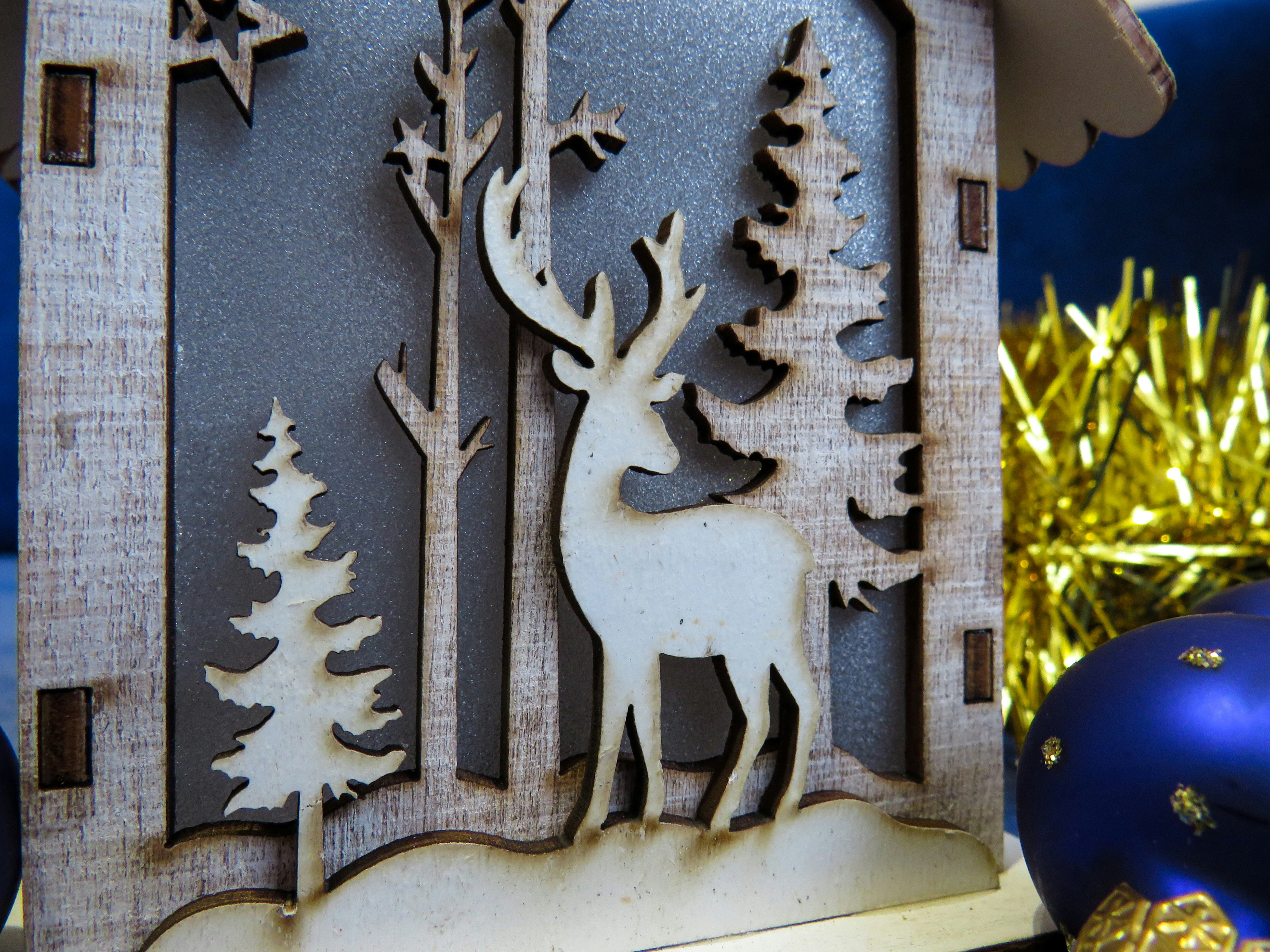 Intricate wooden decoration featuring a stag amidst stylized trees, set against a shimmering backdrop. The scene evokes a serene winter atmosphere.