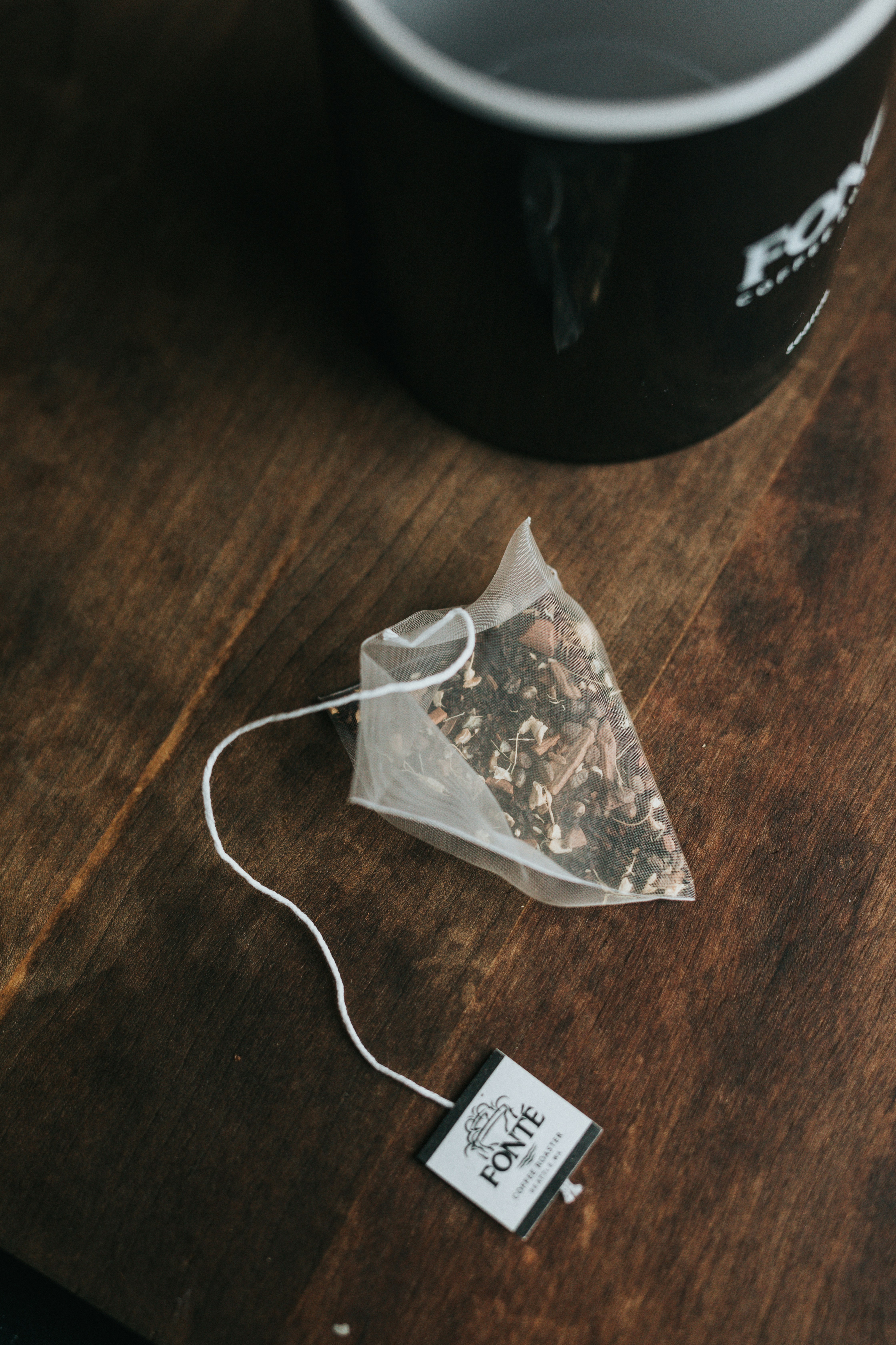 Tea bag resting on a wooden surface beside a black mug, ready for brewing. The delicate details of the tea leaves are visible through the bag.
