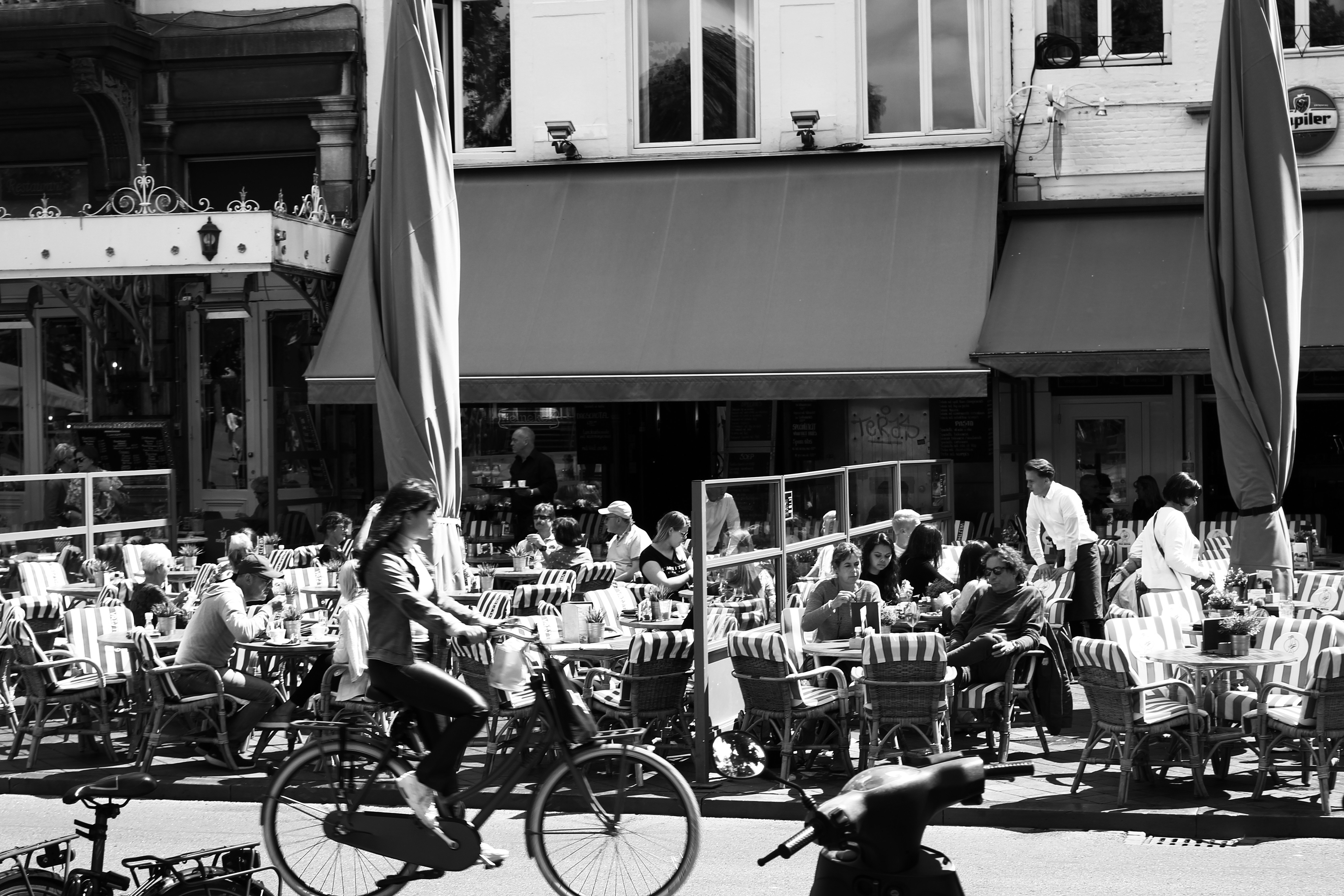 Cyclist passing bustling outdoor café in a lively city street.