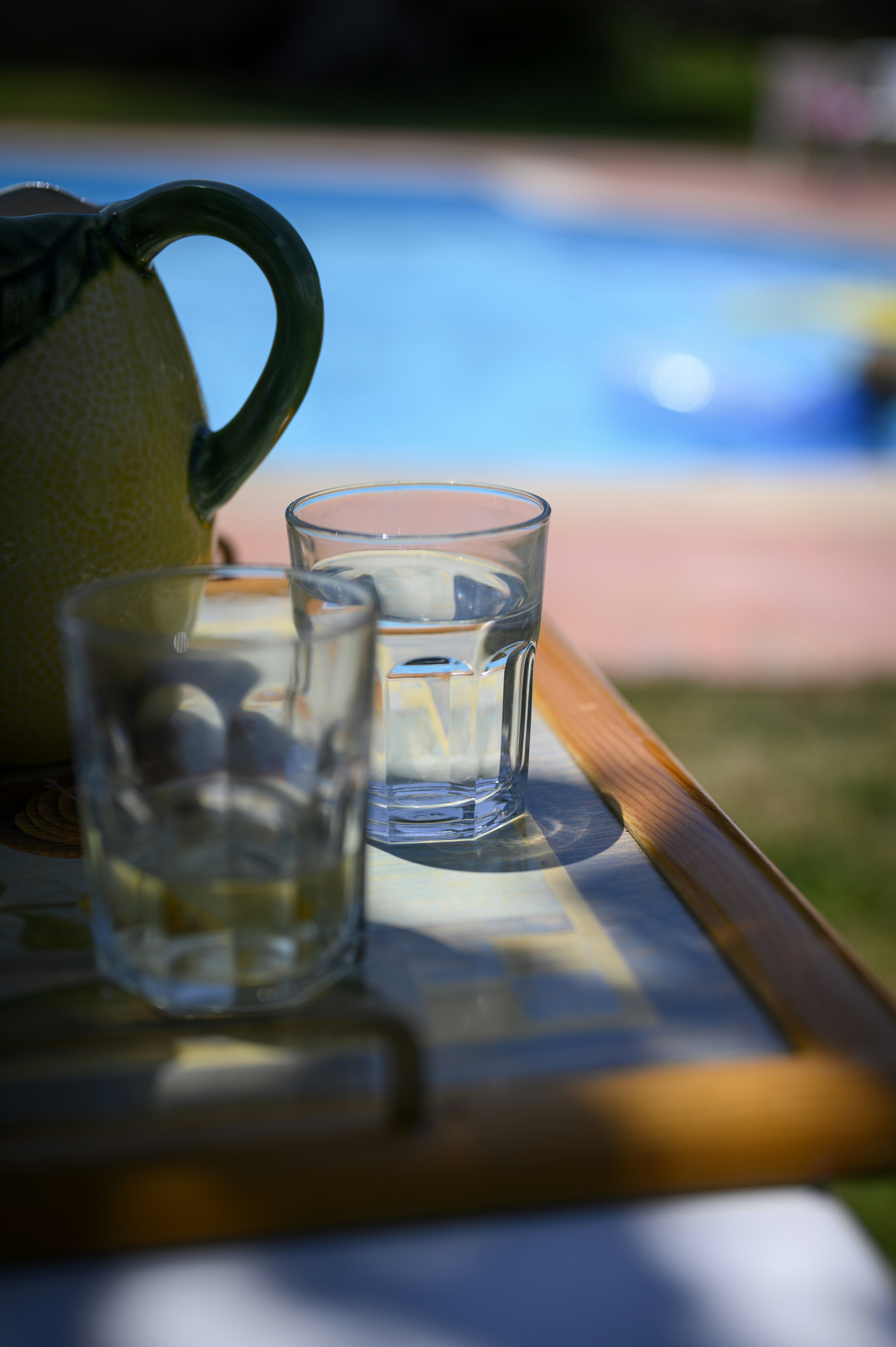 Glass pitcher and two water glasses rest on a wooden tray beside a sunlit pool, with soft background bokeh.