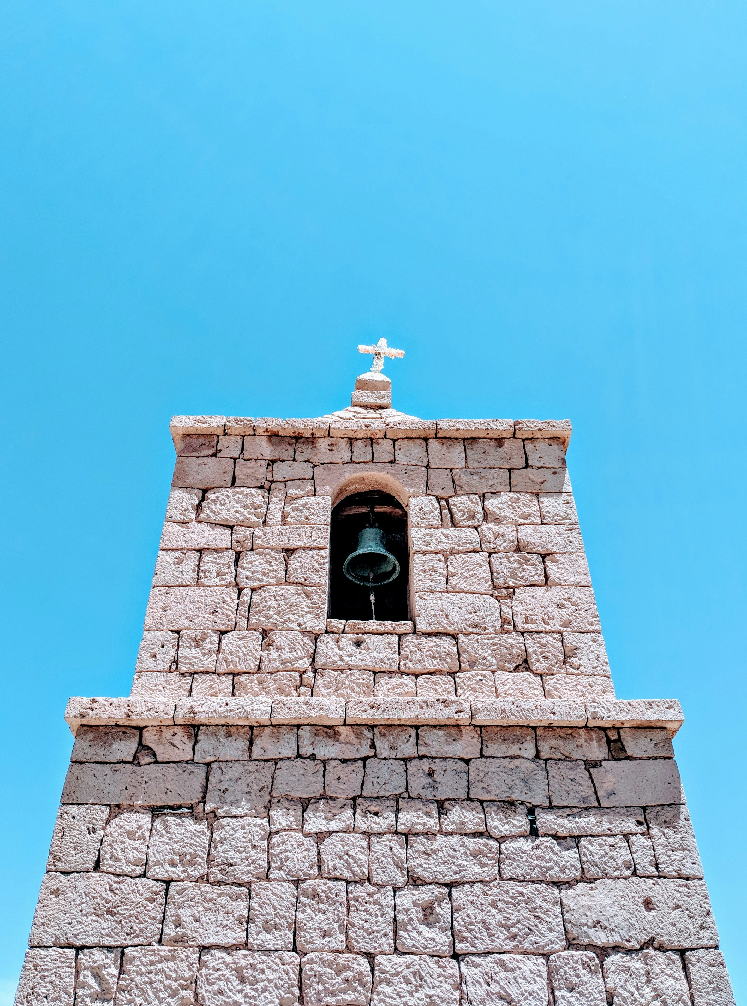 Historic stone bell tower against a clear blue sky, featuring a prominent cross at its peak. The bell hangs in the archway, suggesting a connection to the past.
