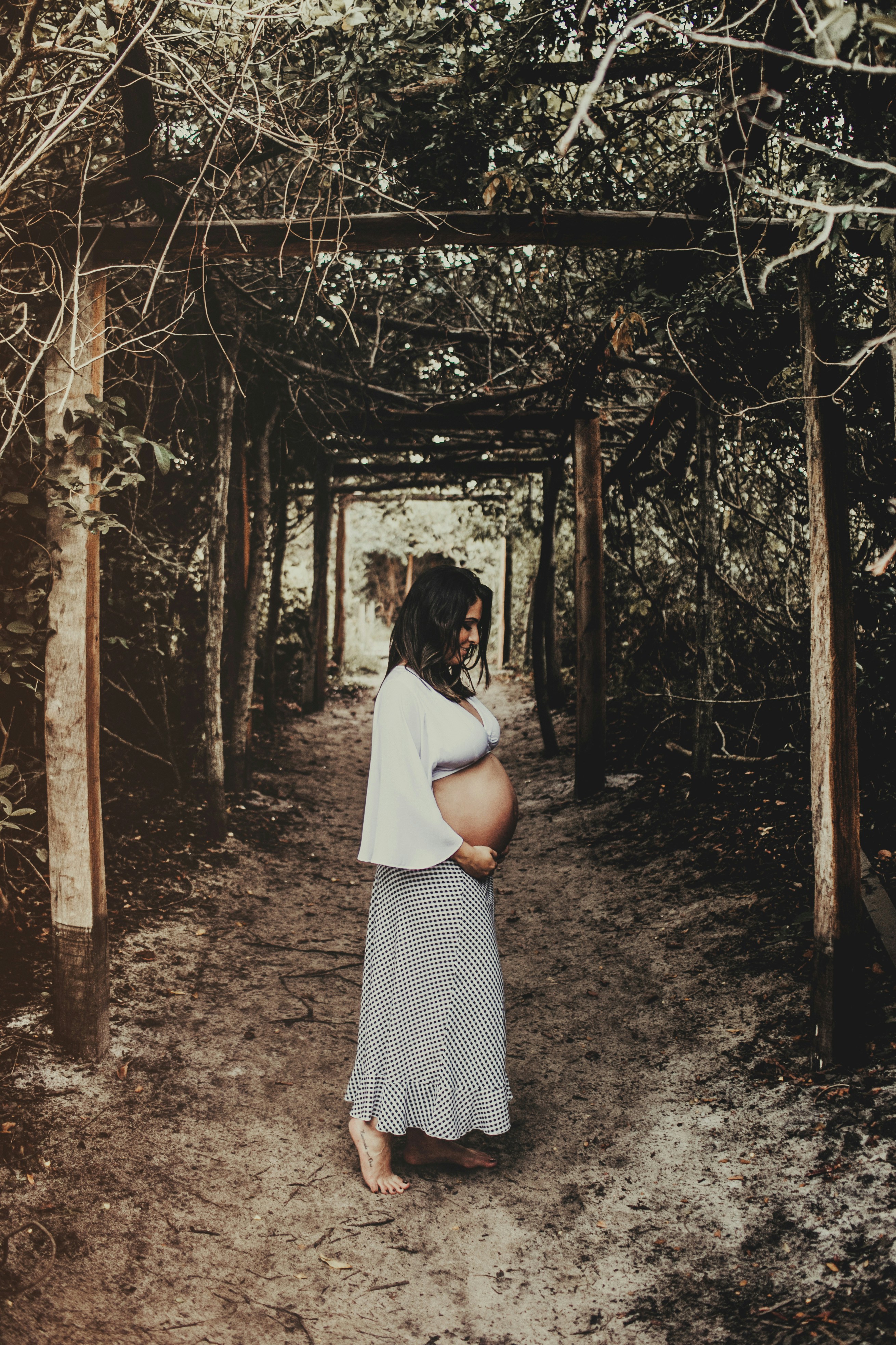 Pregnant woman standing barefoot on a sandy path, cradling her belly under a wooden archway adorned with greenery.