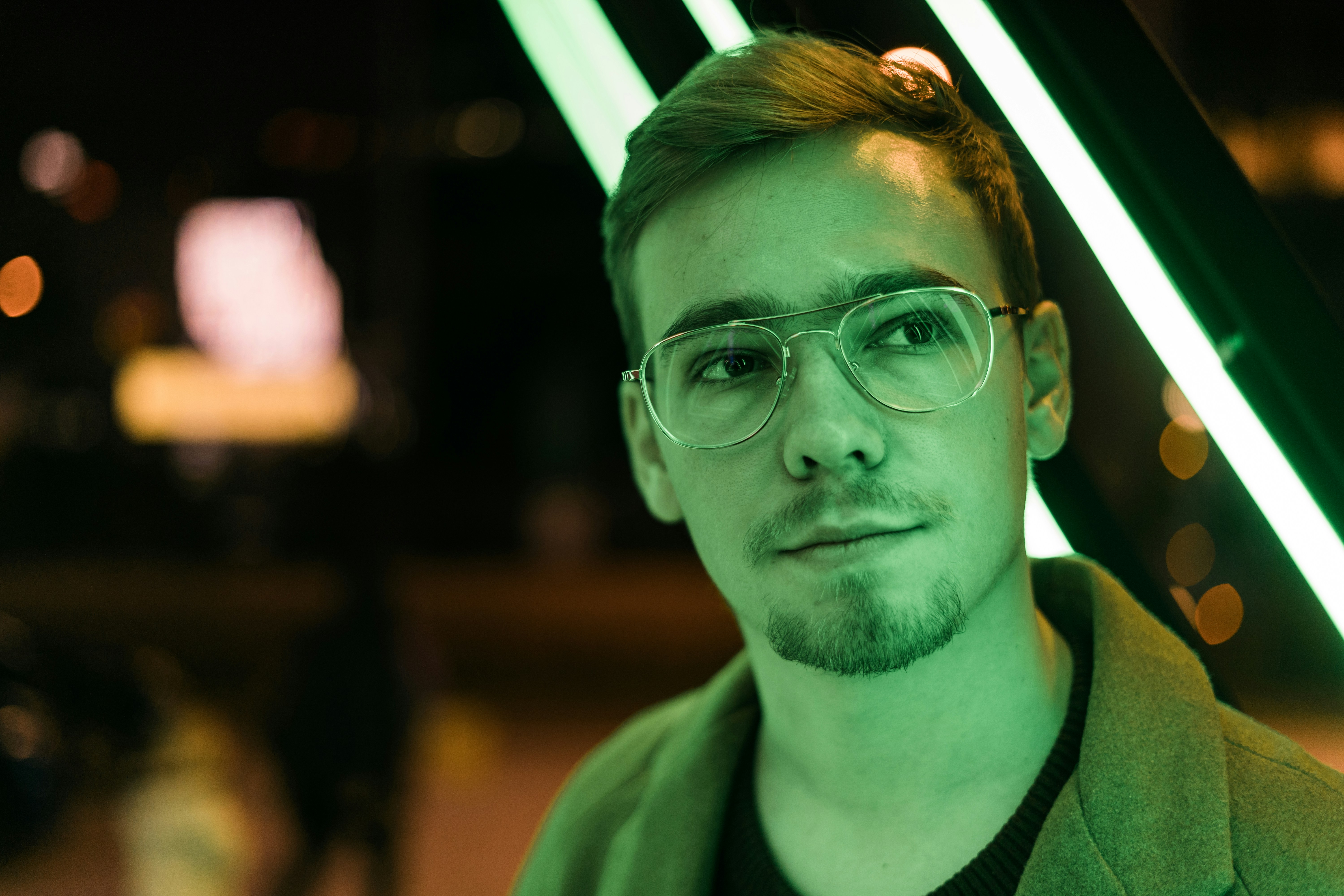 Man with glasses illuminated by green neon lights, standing against a blurred city backdrop at night.