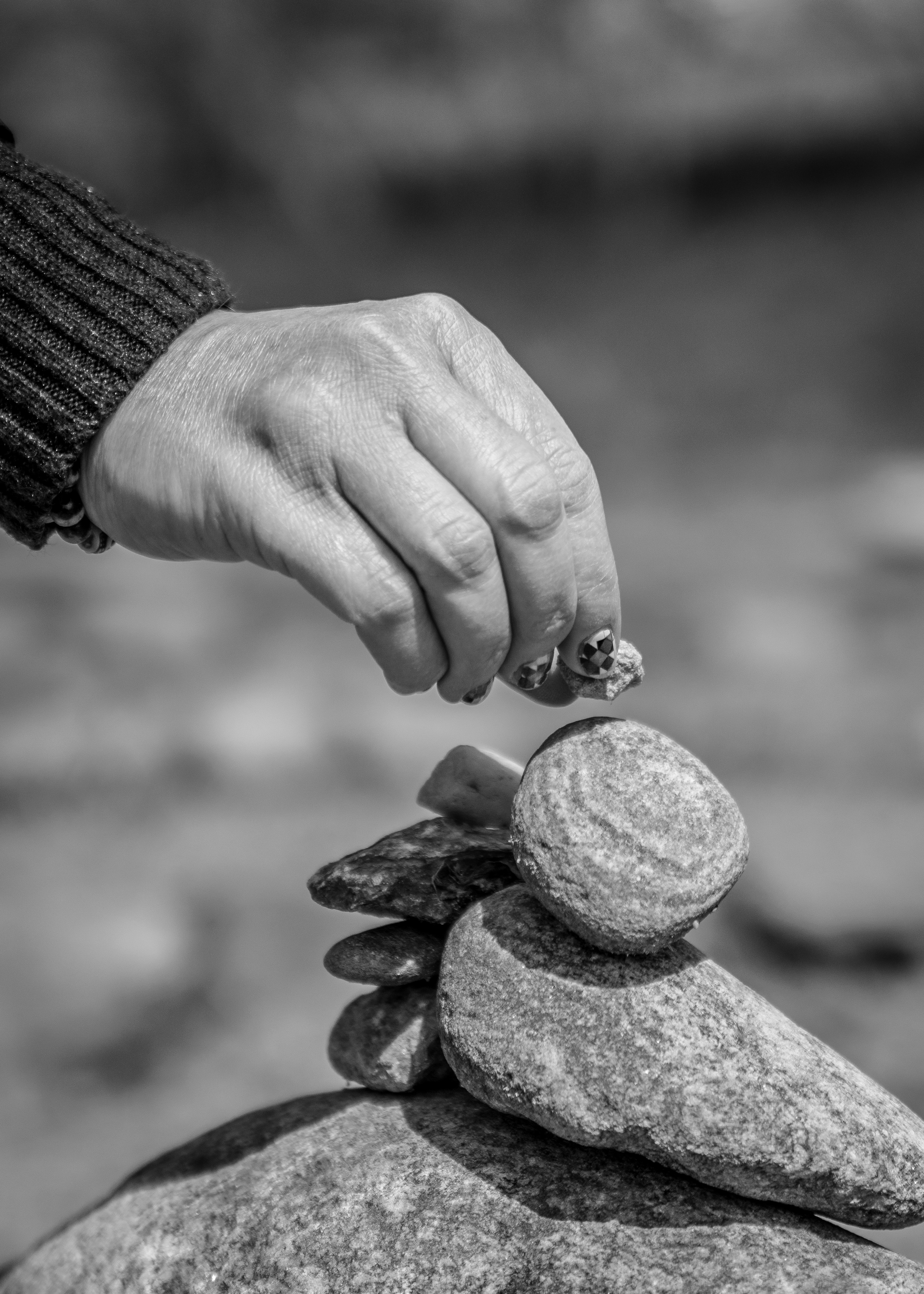 A hand delicately places a stone atop a carefully balanced stack of rocks, showcasing artistry in nature. The monochrome tones emphasize the textures and shapes of the stones.