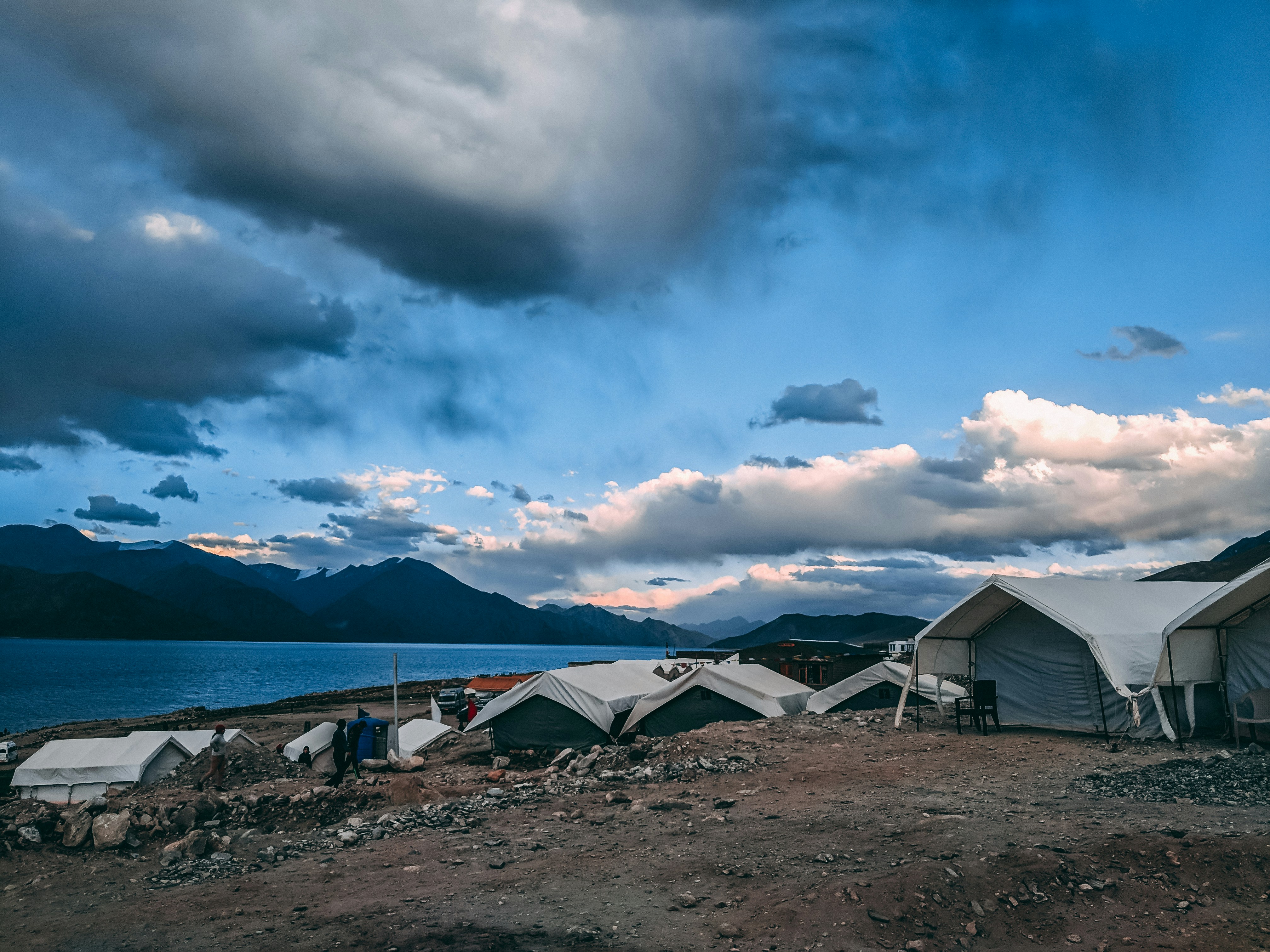 Tents arranged along a lakeshore with mountains in the background under a dramatic sky. The scene captures the essence of outdoor adventure and tranquility.