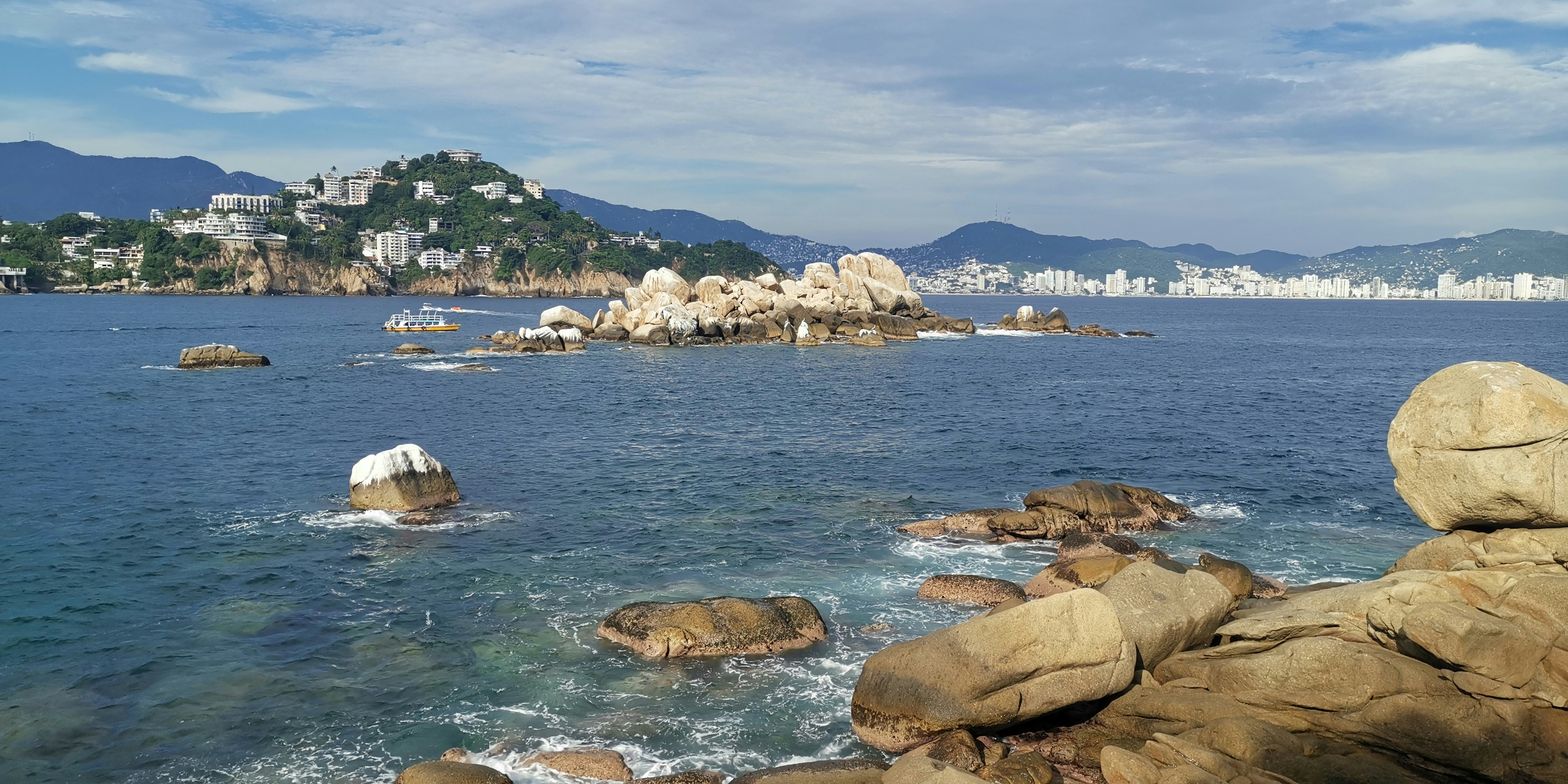 Coastal landscape with weathered foreground rocks, calm blue sea, and a distant hillside town perched above the bay.