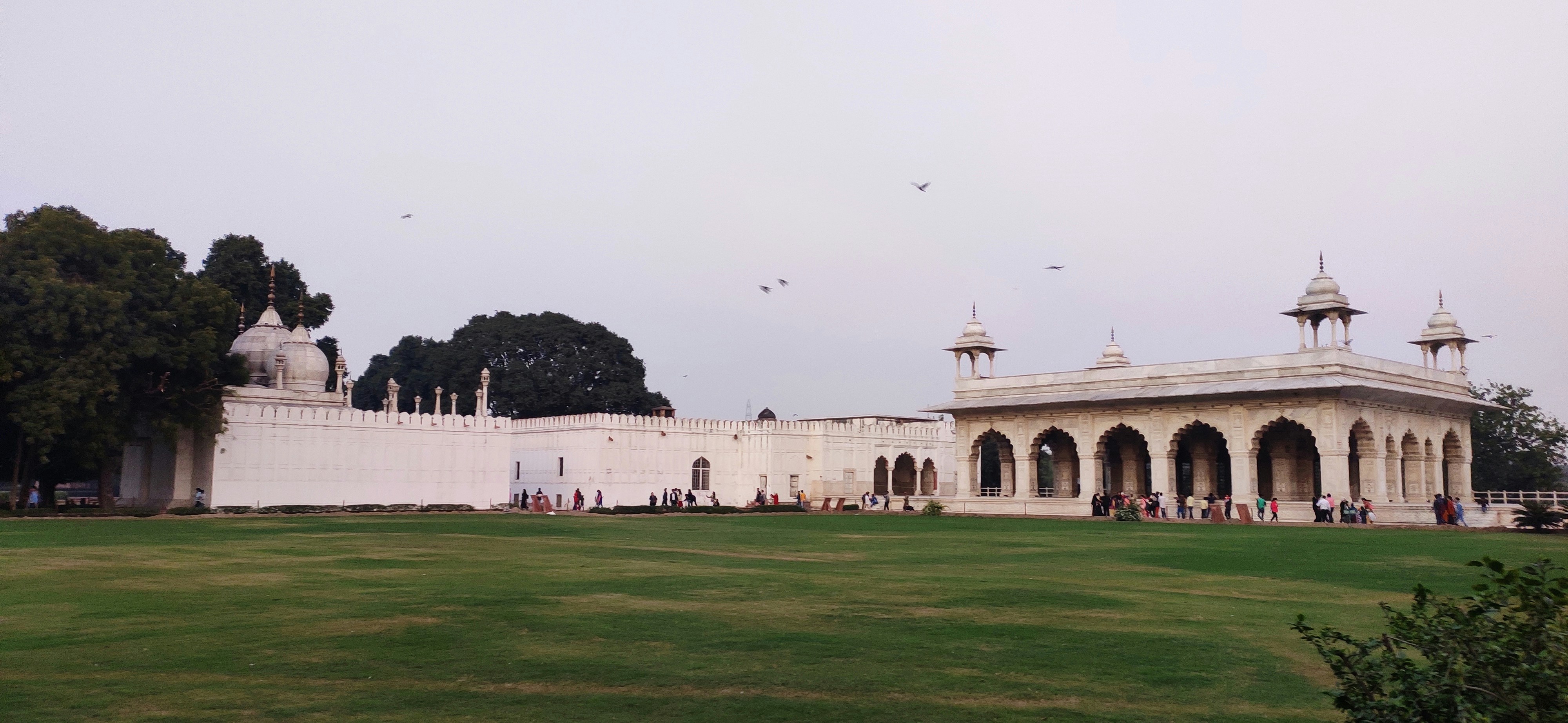 An expansive view of a historic architectural complex featuring intricate domes and arches, surrounded by lush green lawns and a gathering of visitors.