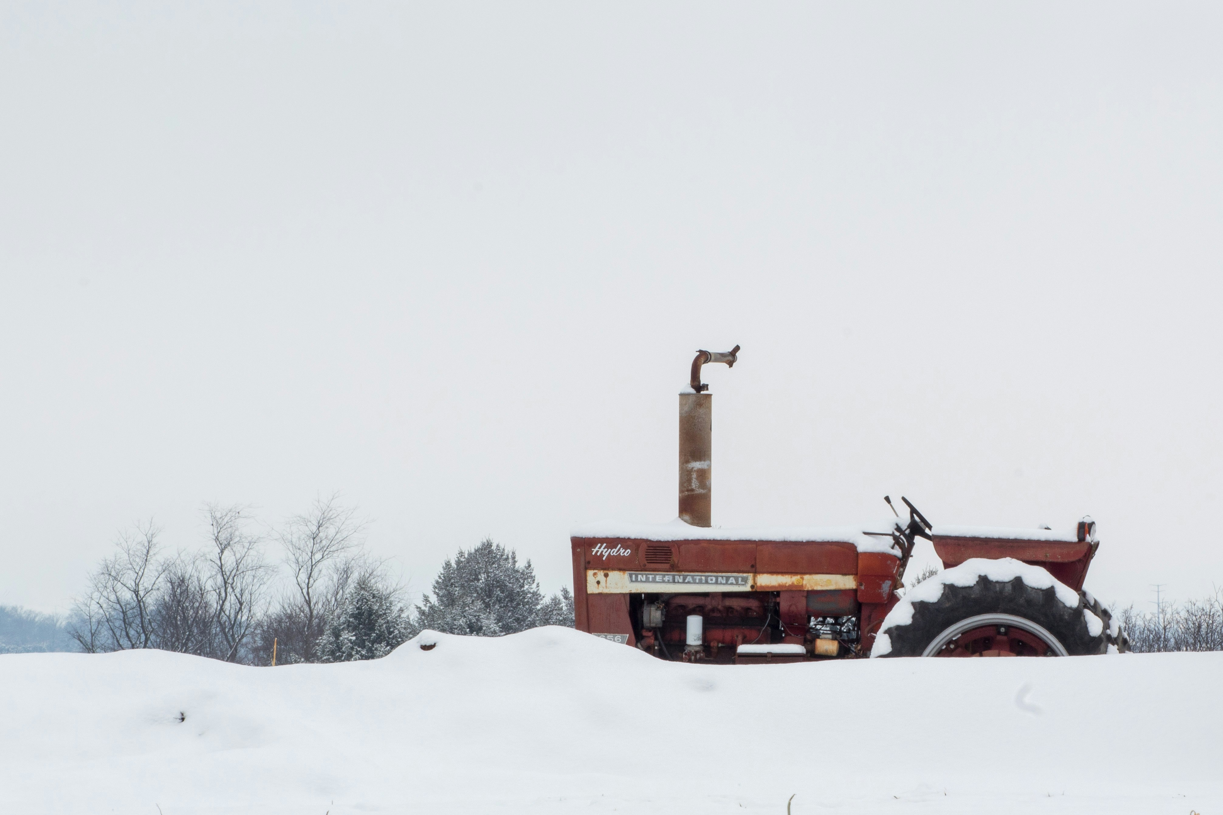 Vintage tractor partially buried in snow, surrounded by bare trees under a gray sky.