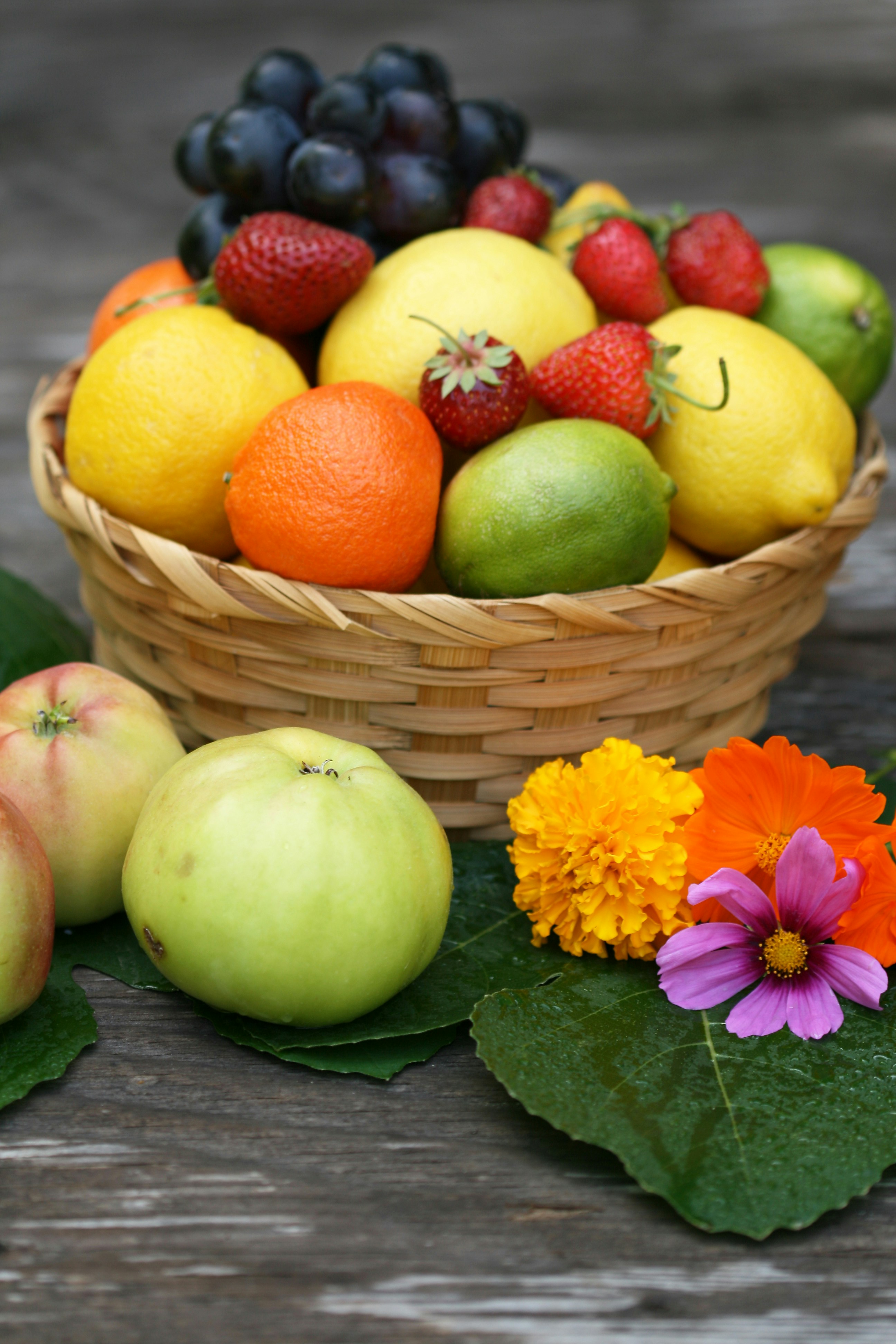 A woven basket brimming with a colorful assortment of fruits, including lemons, strawberries, and grapes, accompanied by vibrant flowers and green leaves.