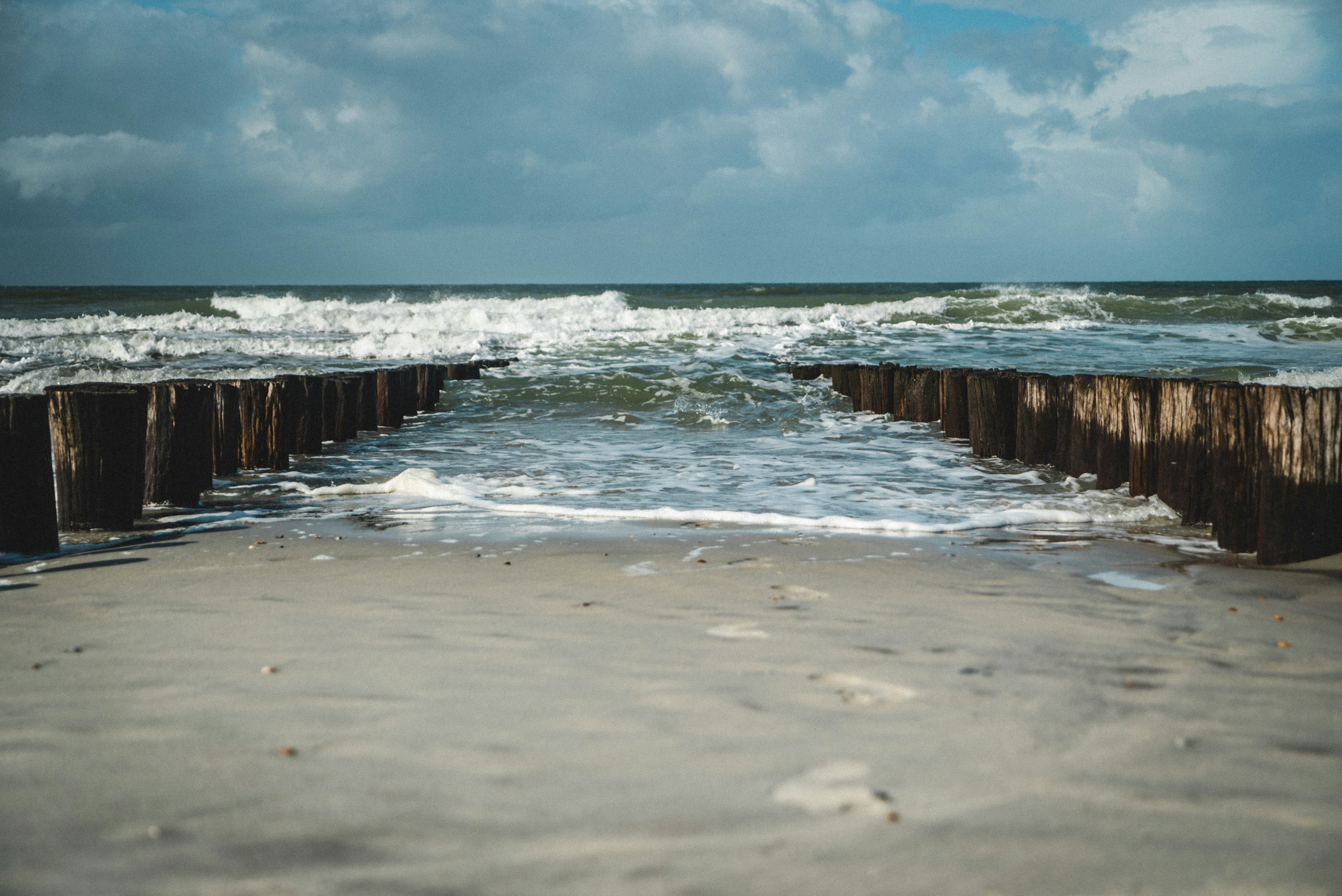 Weathered wooden posts jut into the waves, guiding the eye toward the horizon where sea meets sky. The dynamic interplay of water and sand creates a serene coastal atmosphere.