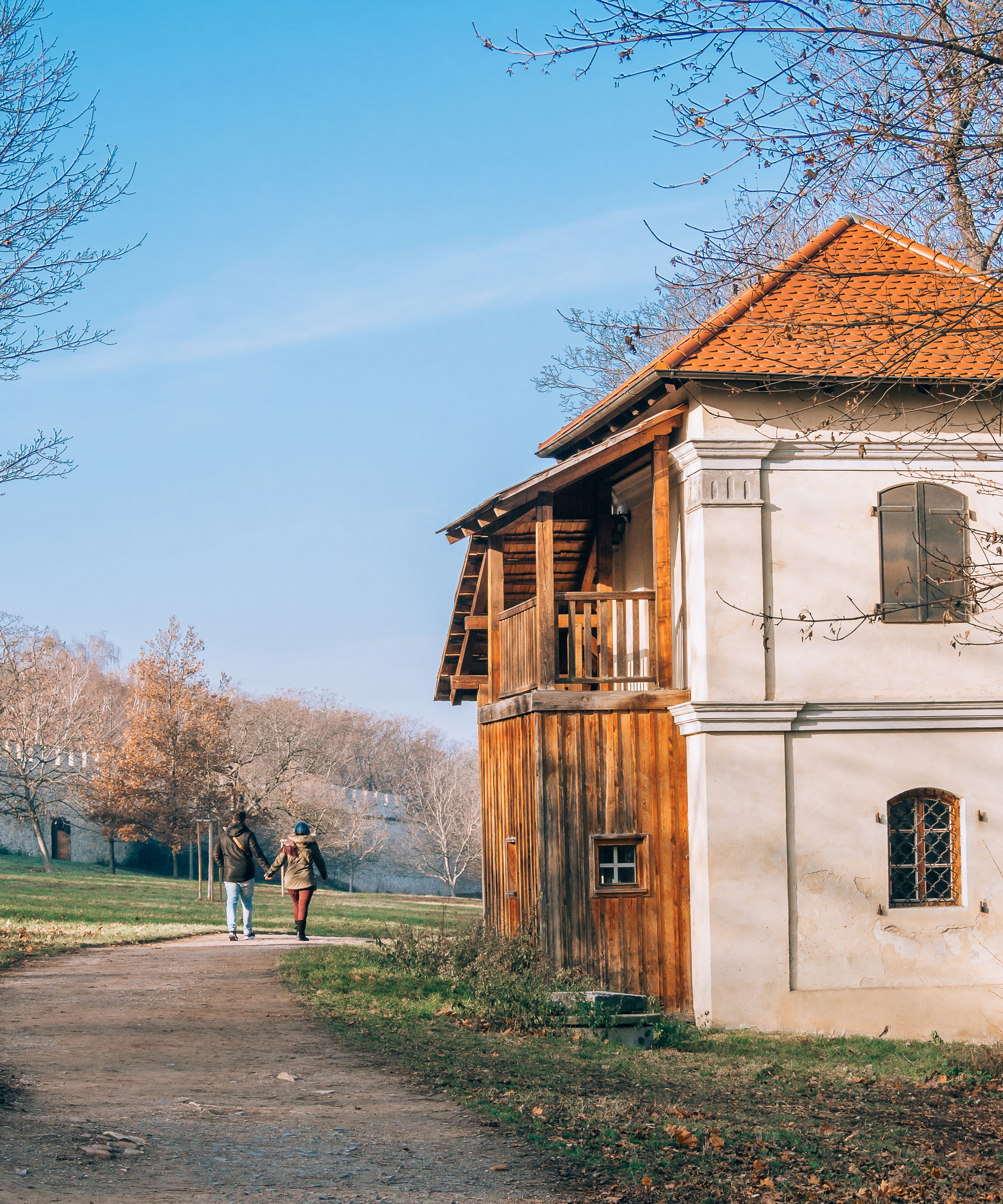 Two friends walking along a path beside a rustic wooden house, framed by autumn trees under a clear blue sky.