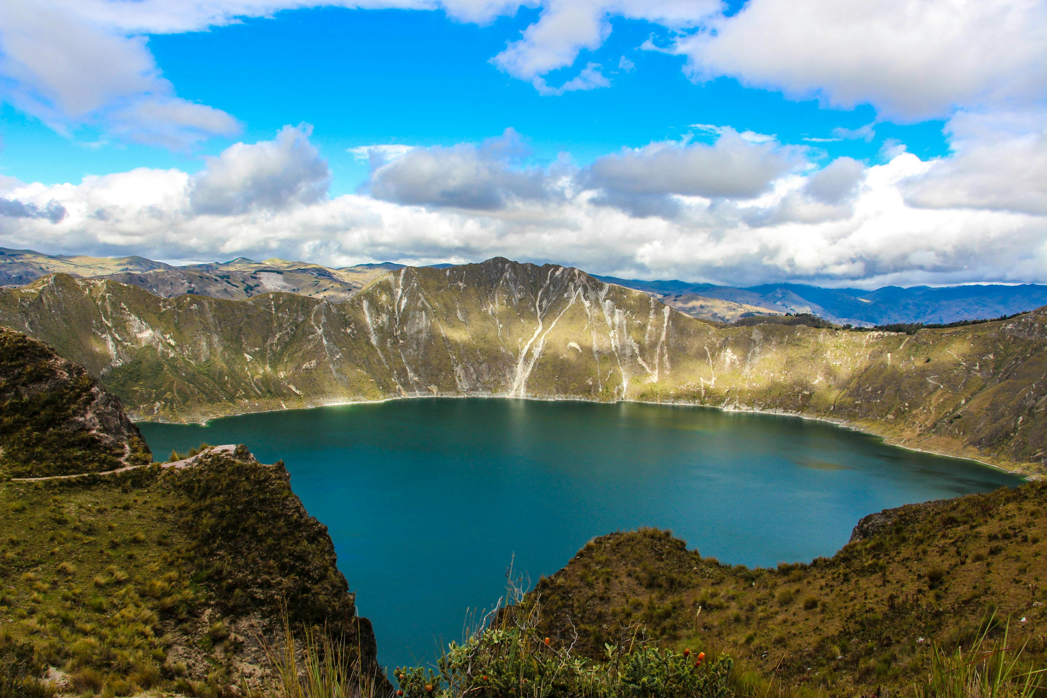 Vibrant blue lake nestled within a volcanic crater, surrounded by rugged mountains and lush greenery under a partly cloudy sky.