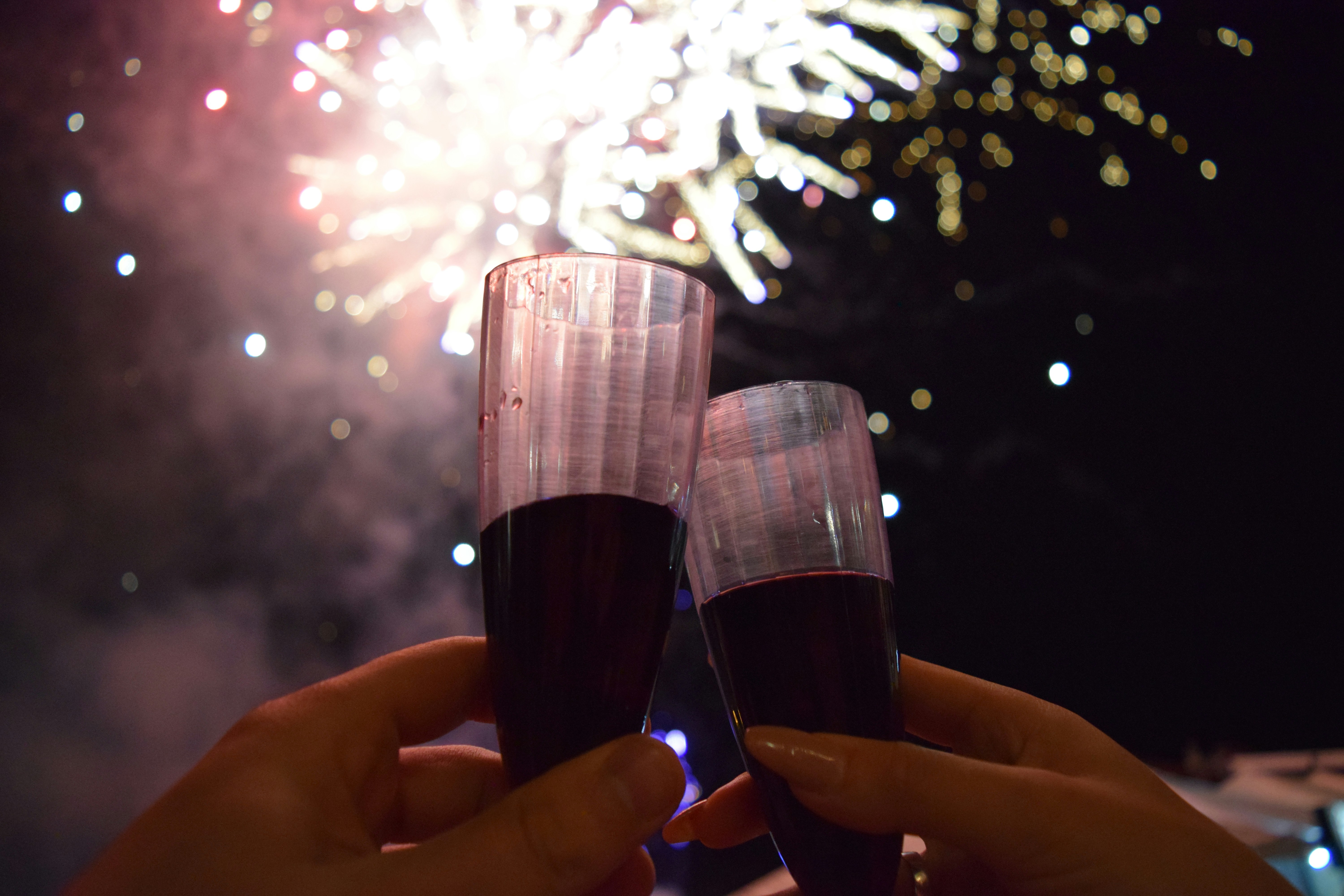Two hands clink glasses of red wine against a backdrop of vibrant fireworks in the night sky.