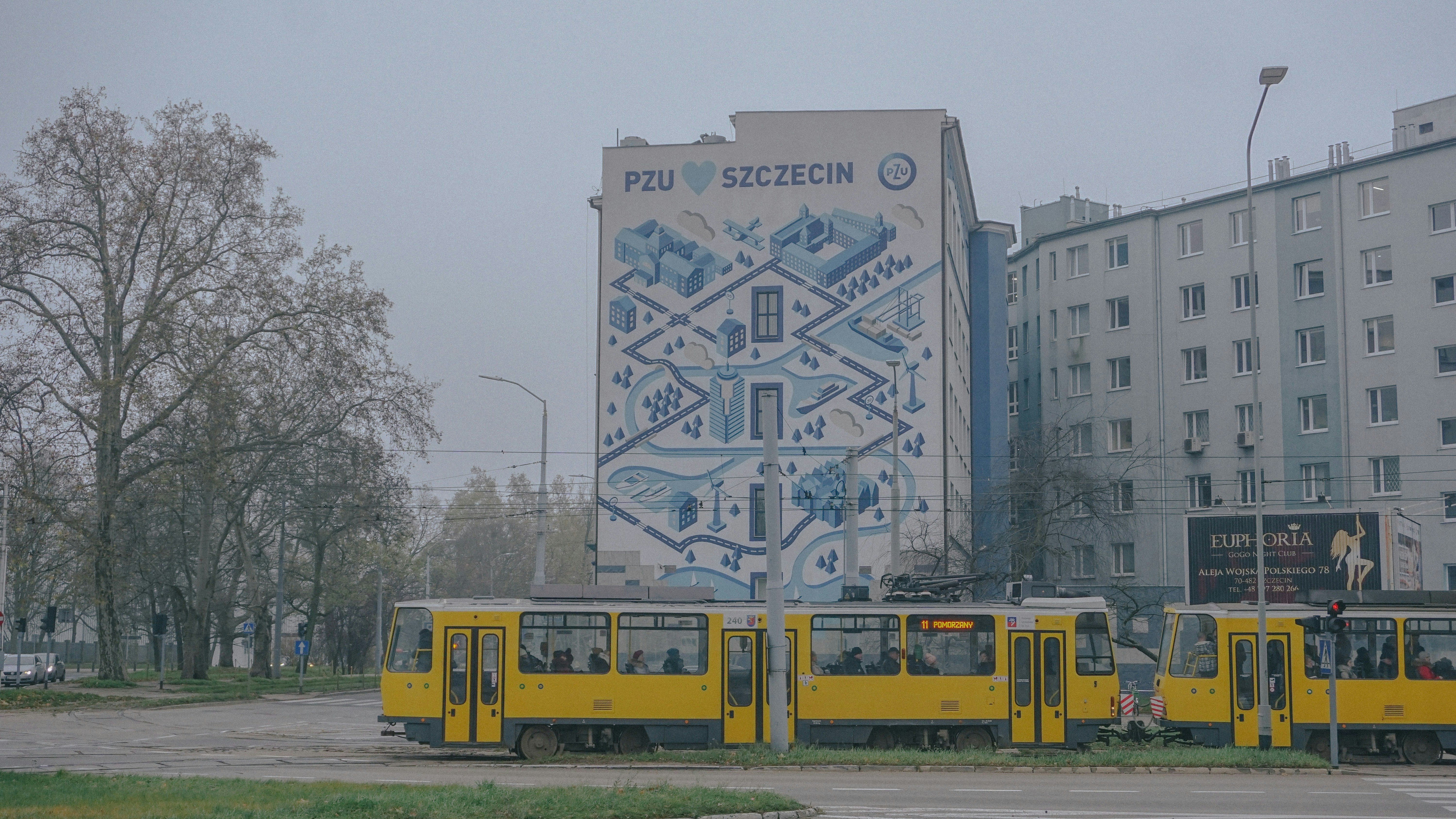 A vibrant mural depicting a cityscape on a building, with yellow trams parked in the foreground under a grey sky.