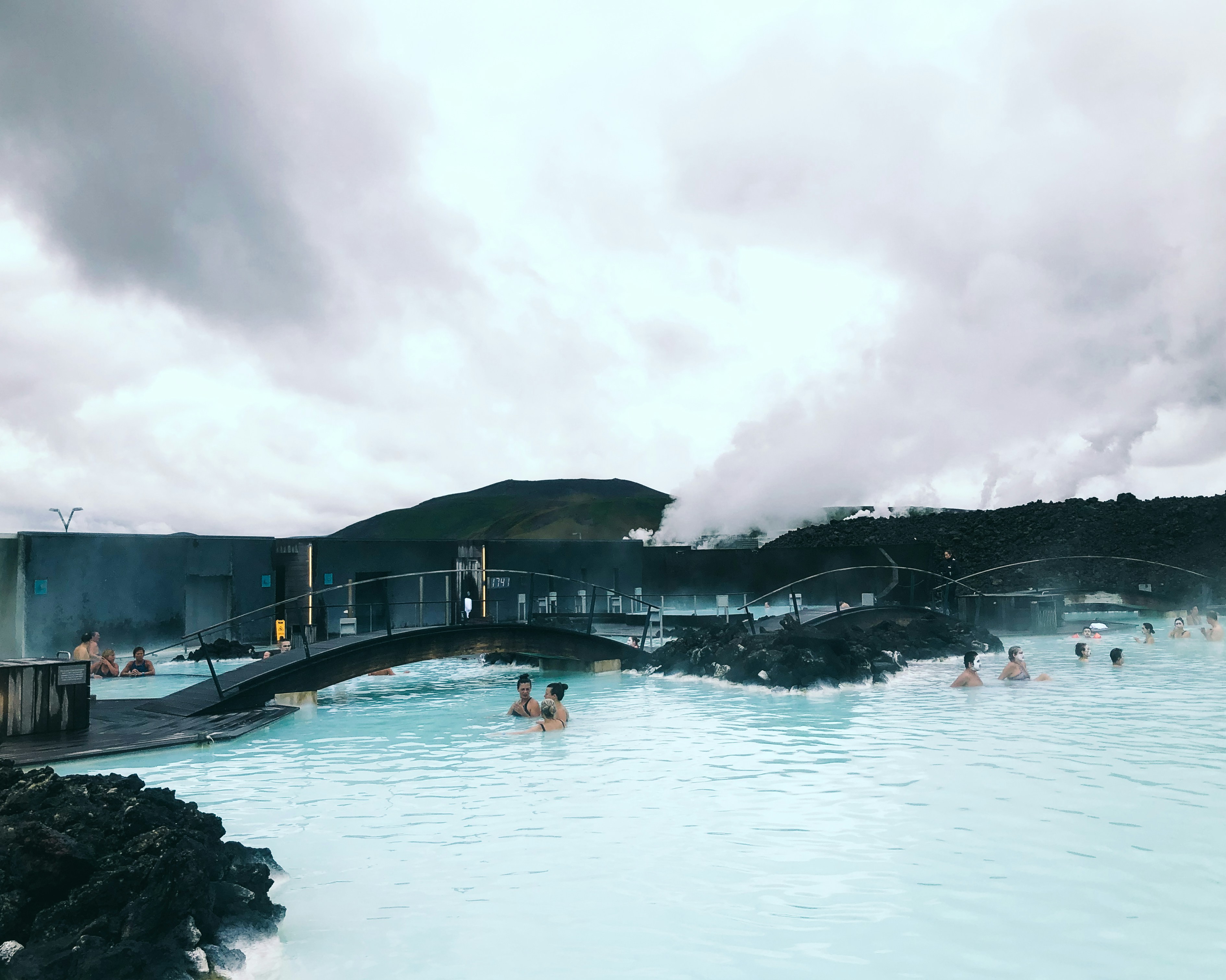 Visitors relax in a geothermal spa surrounded by steam and rocky formations under a cloudy sky.