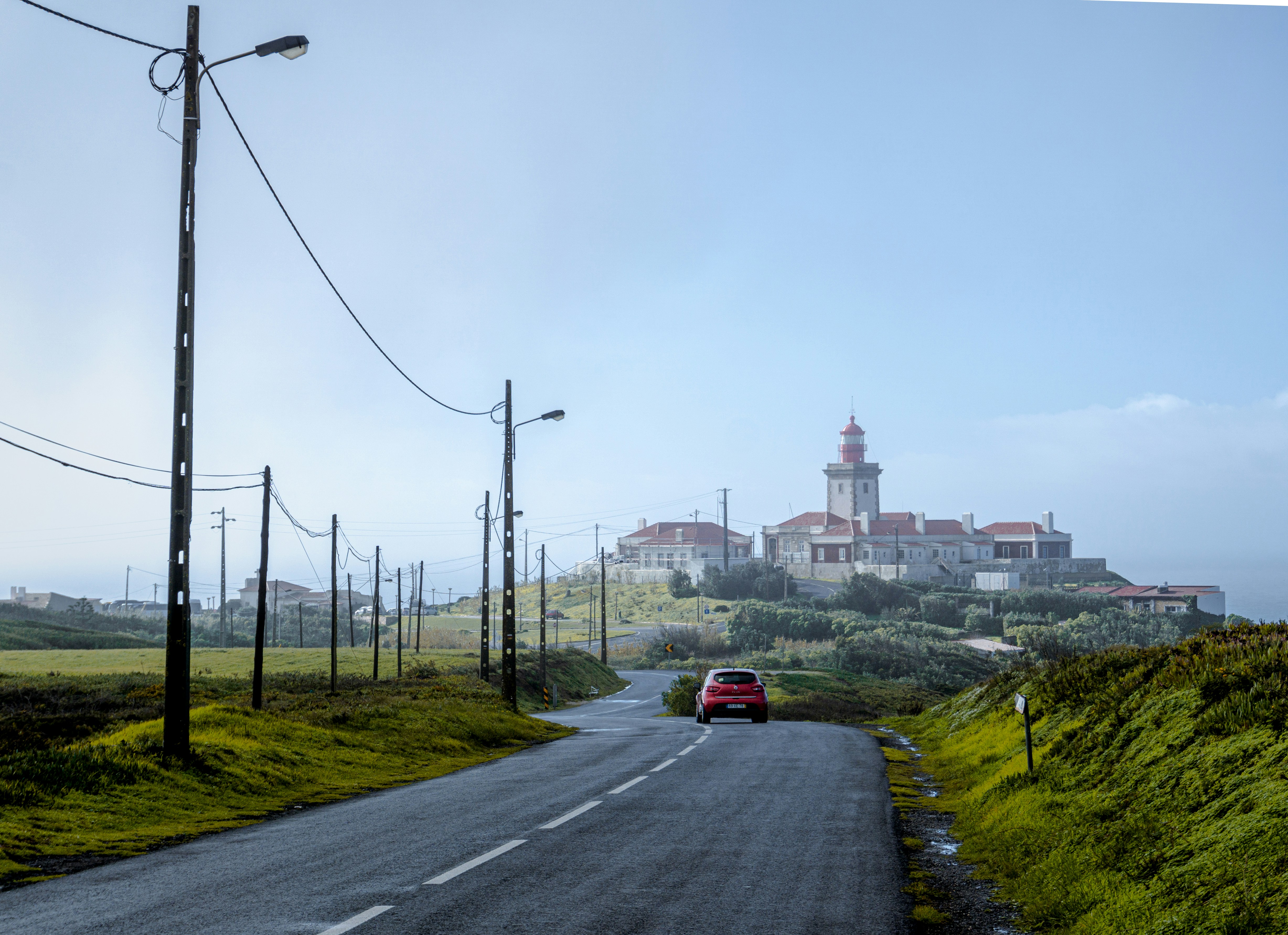 A winding road leads to a lighthouse perched on a hill, surrounded by lush greenery and a misty sky.